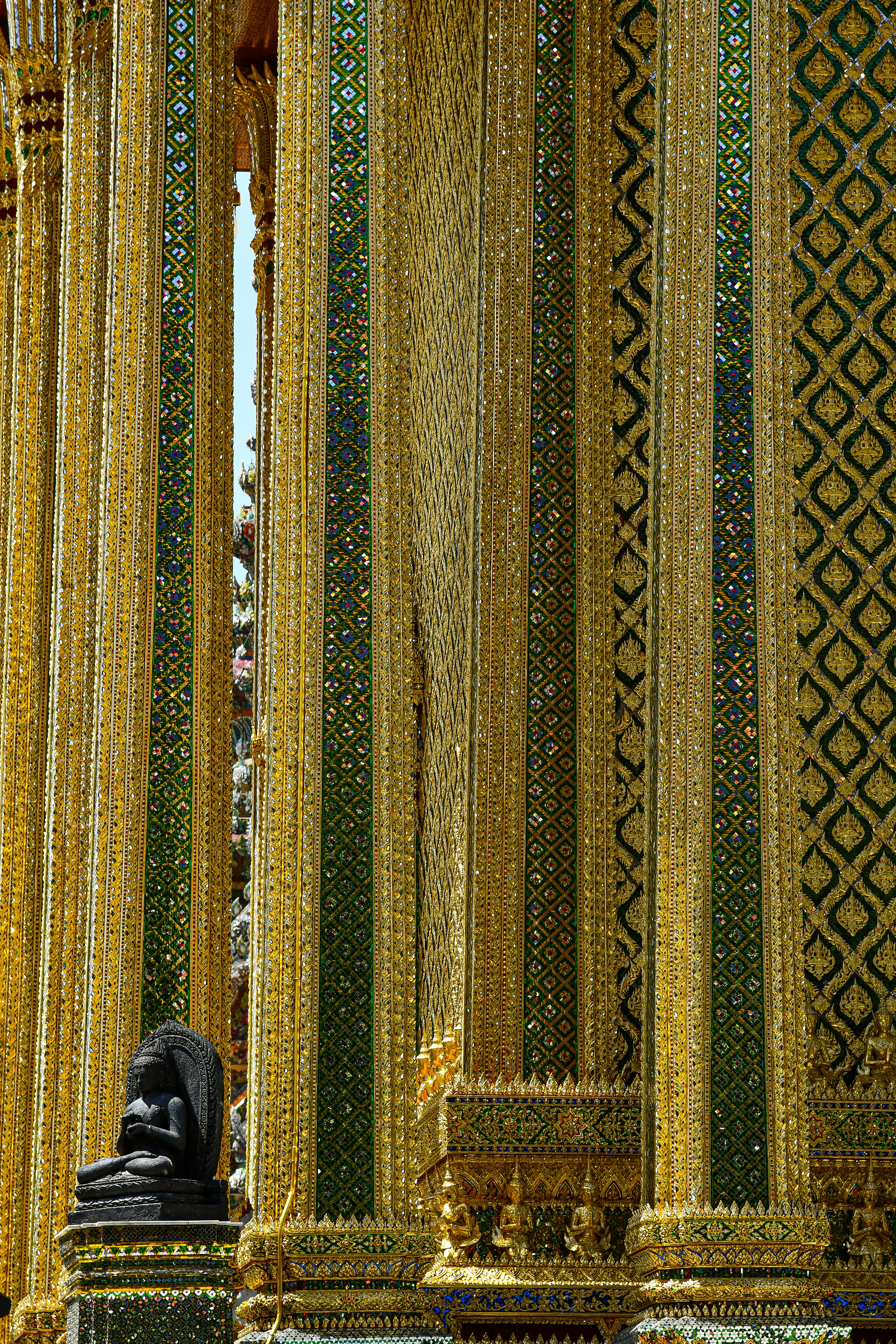 Golden temple columns with intricate green and diamond-patterned inlays frame a small carved sculpture at the base, highlighting shimmering surfaces and ornate craftsmanship.