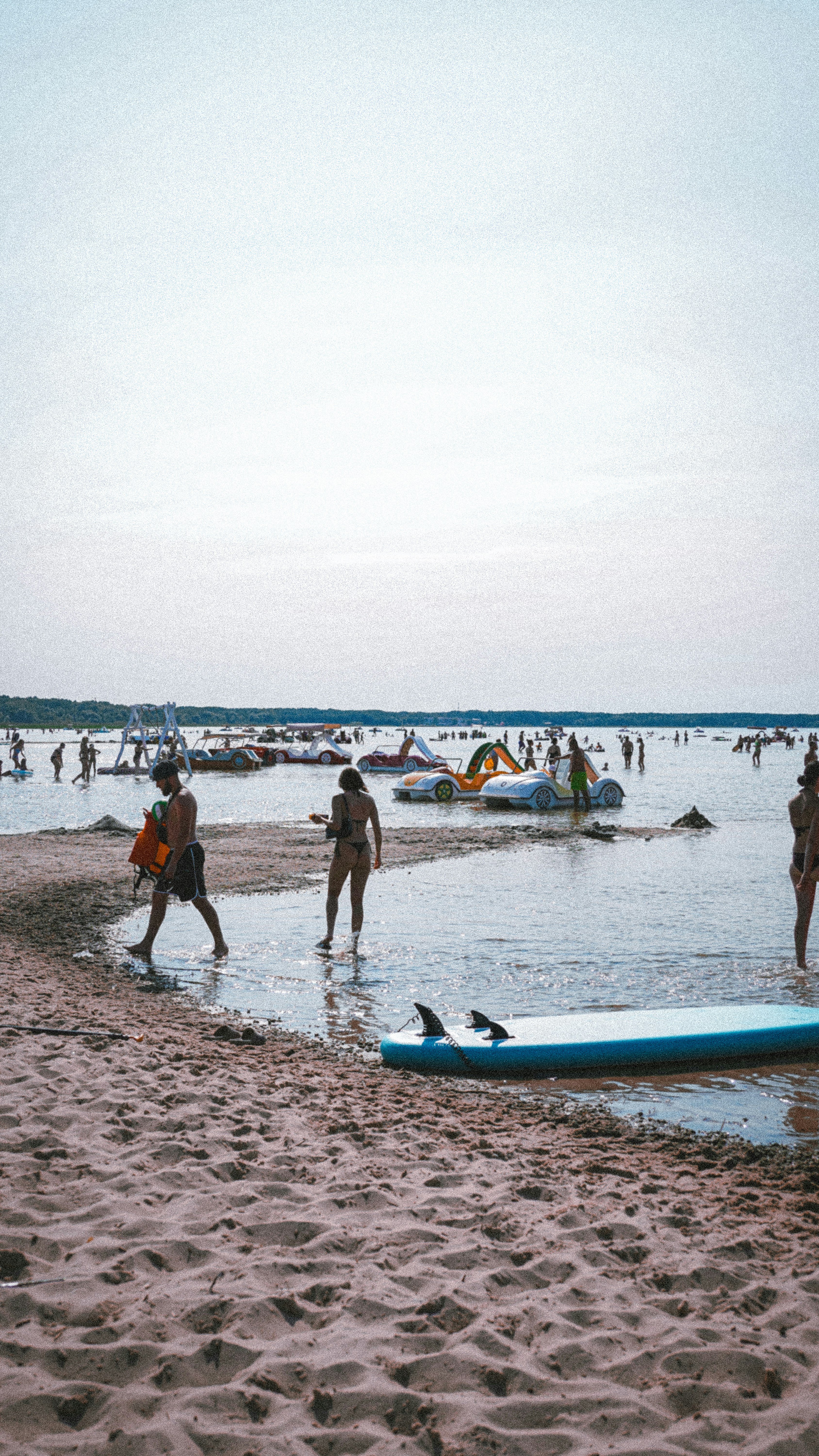 Bright beach scene with people walking along the shore and a blue surfboard in the foreground; boats and groups of swimmers fill the shallow water, with a calm horizon in the distance.