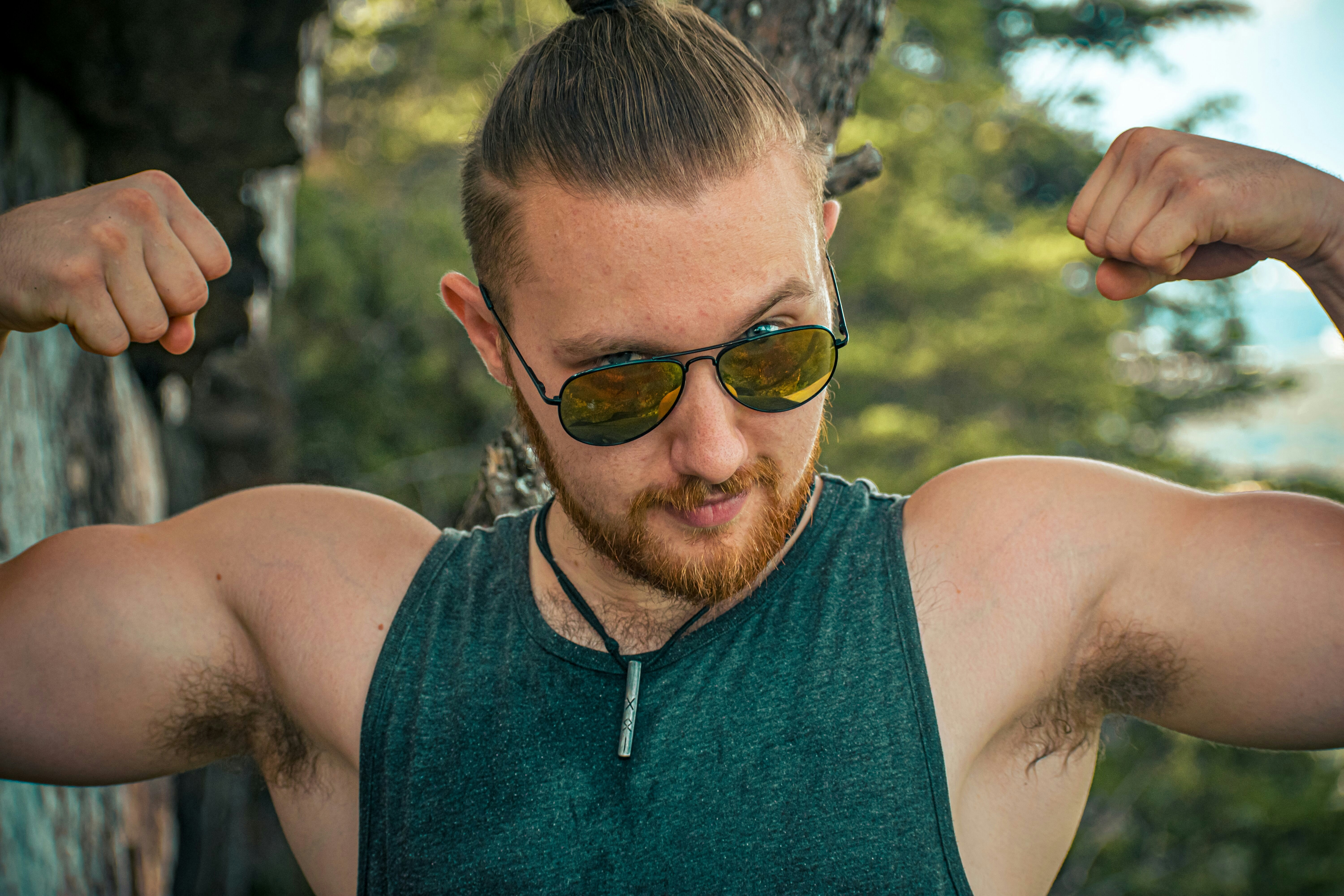 Muscular man wearing sleeveless teal top and sunglasses poses with flexed arms in a sunny outdoor setting.