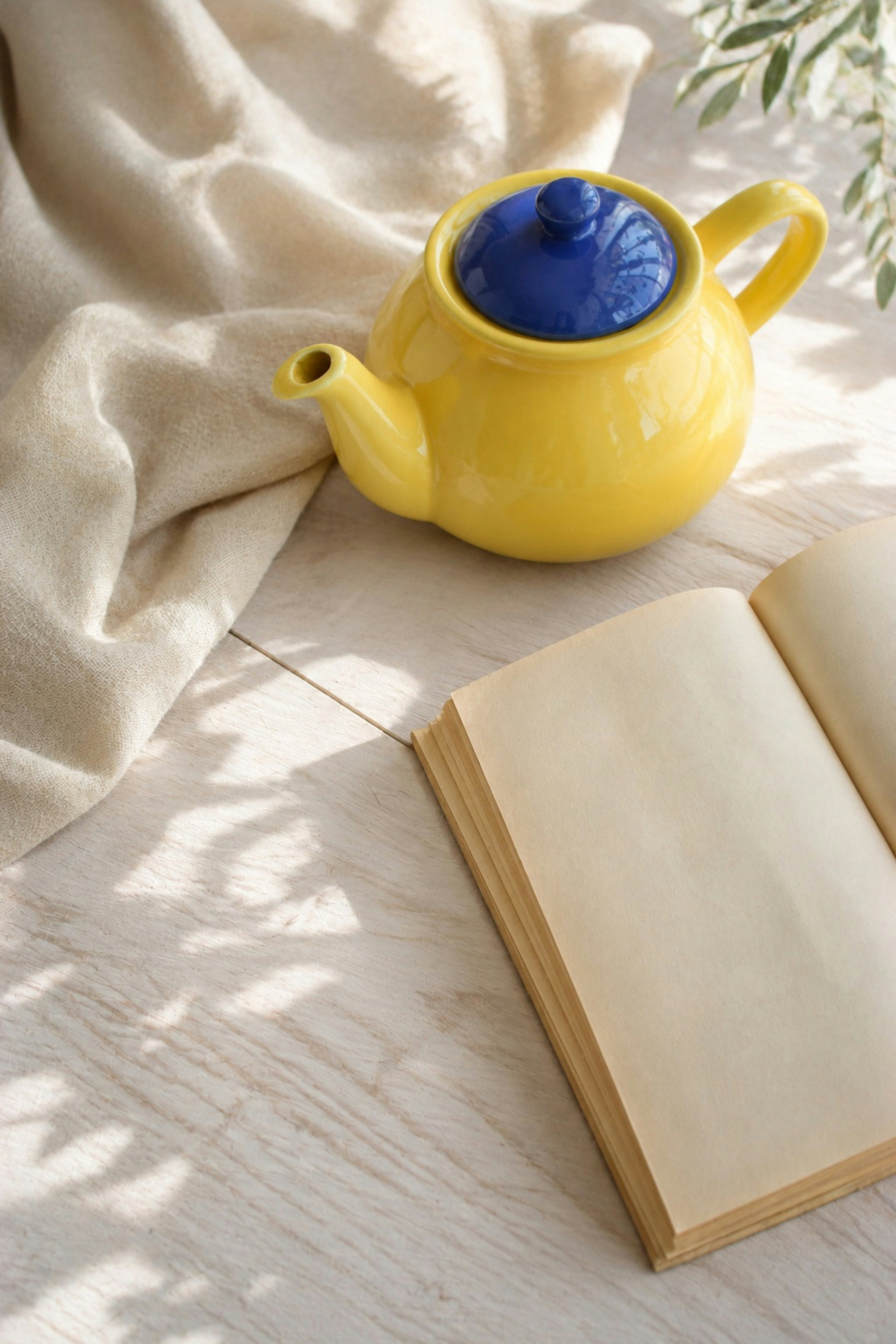 Bright yellow teapot with blue lid sits beside an open notebook on a light wooden surface, with soft fabric and shadow patterns in the background.