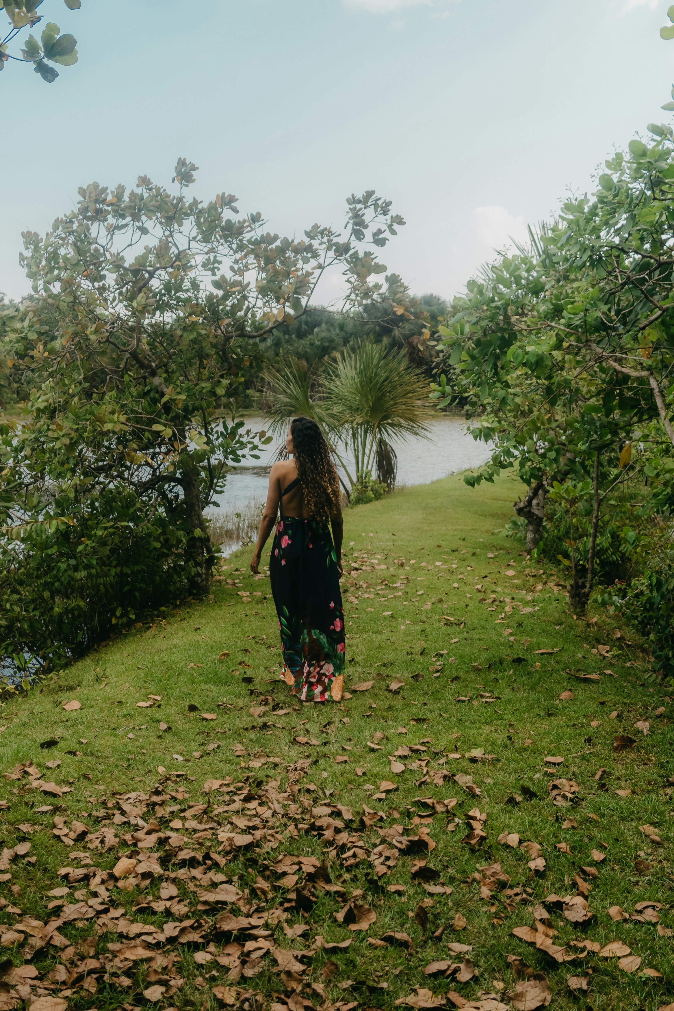 A woman in a long floral dress walks along a grassy path toward a serene lake, surrounded by trees and fallen leaves; natural landscape foreground with a soft, sunlit atmosphere. (photograph)