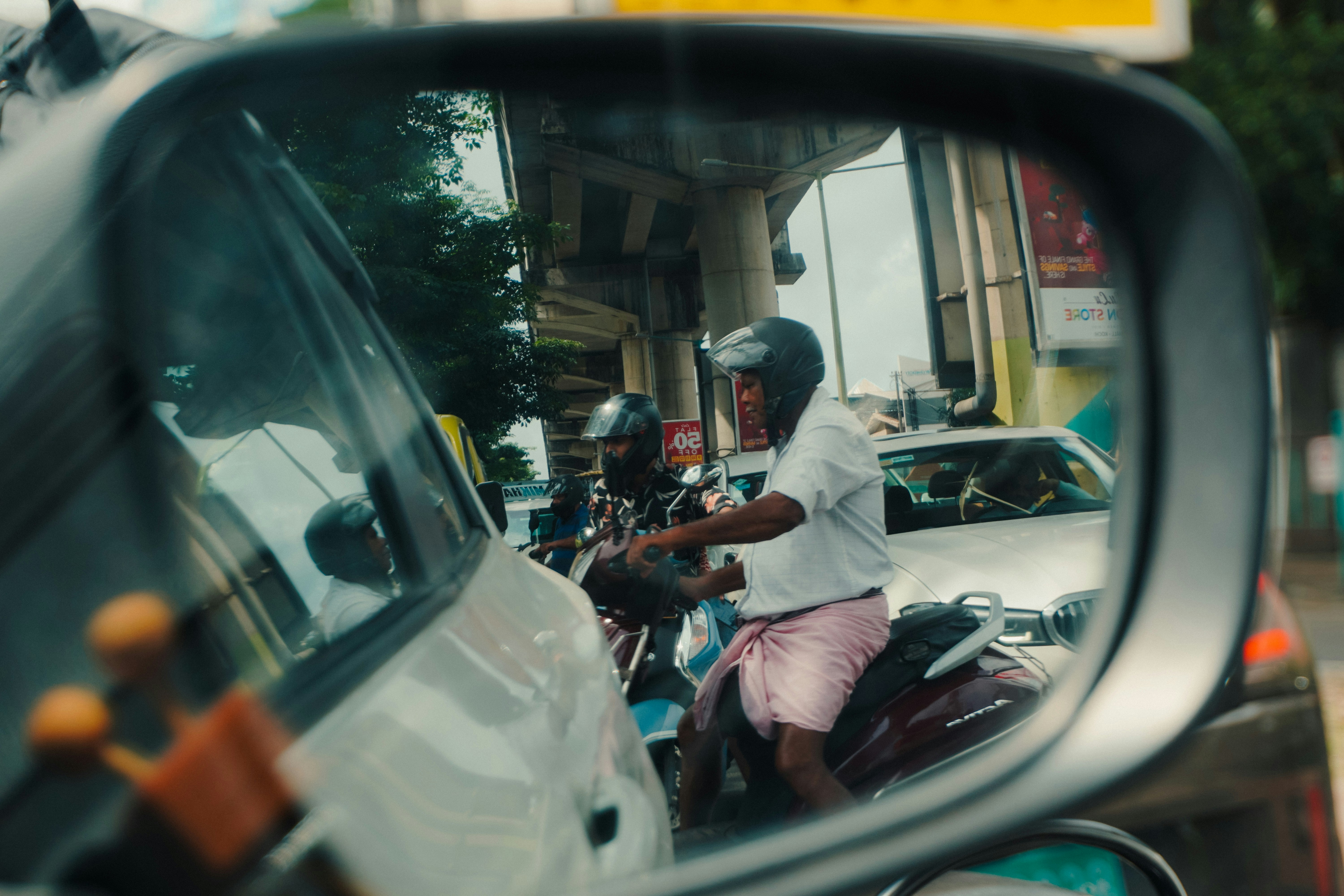 Motorcyclist and nearby vehicles reflected in a car’s side mirror, capturing a busy street scene with shallow depth and motion. This is a candid, photojournalistic moment within an urban environment.