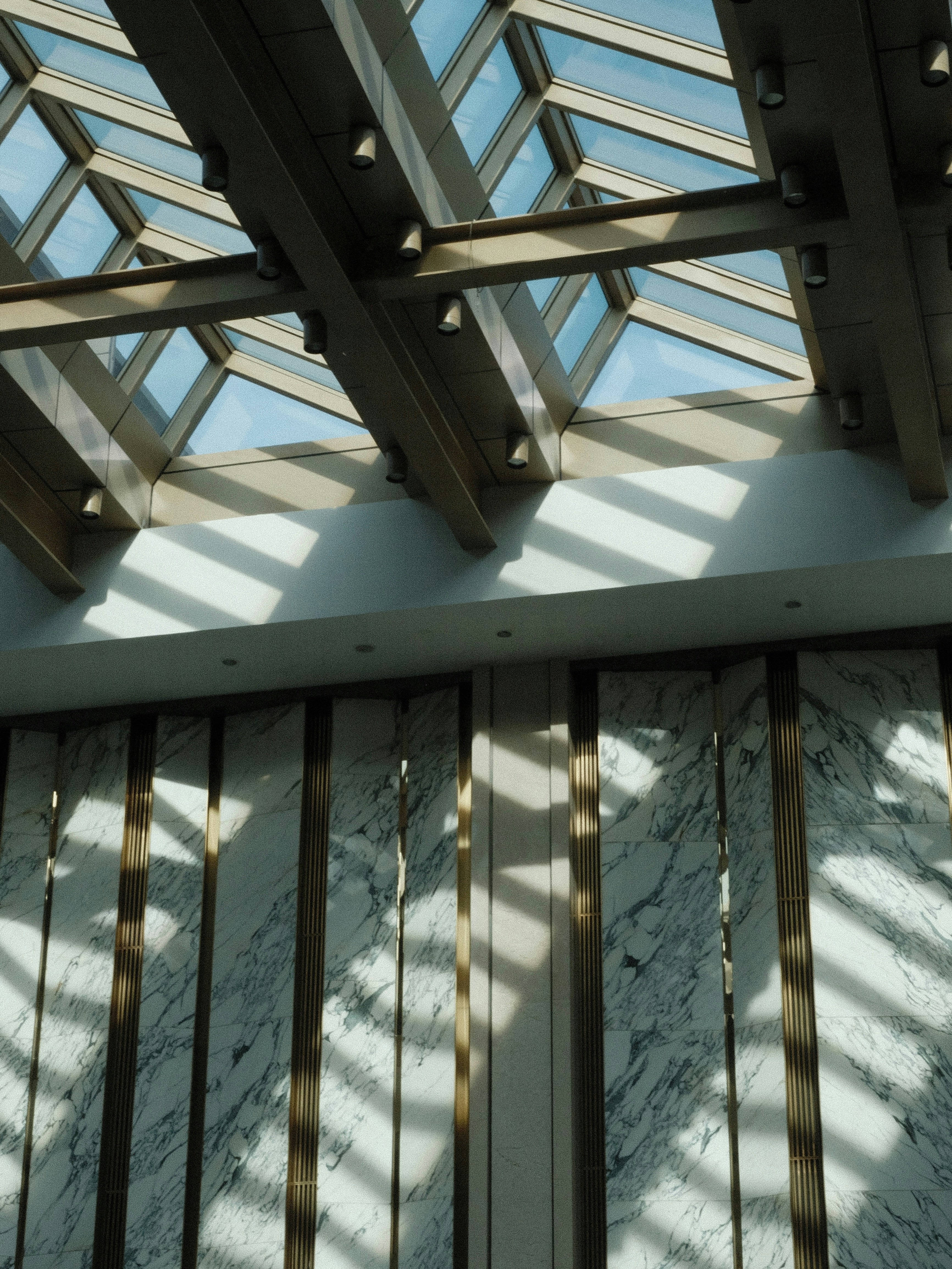 Angular glass skylights and gilded columns cast diagonal shadows over marble walls, revealing architectural rhythm and light play.