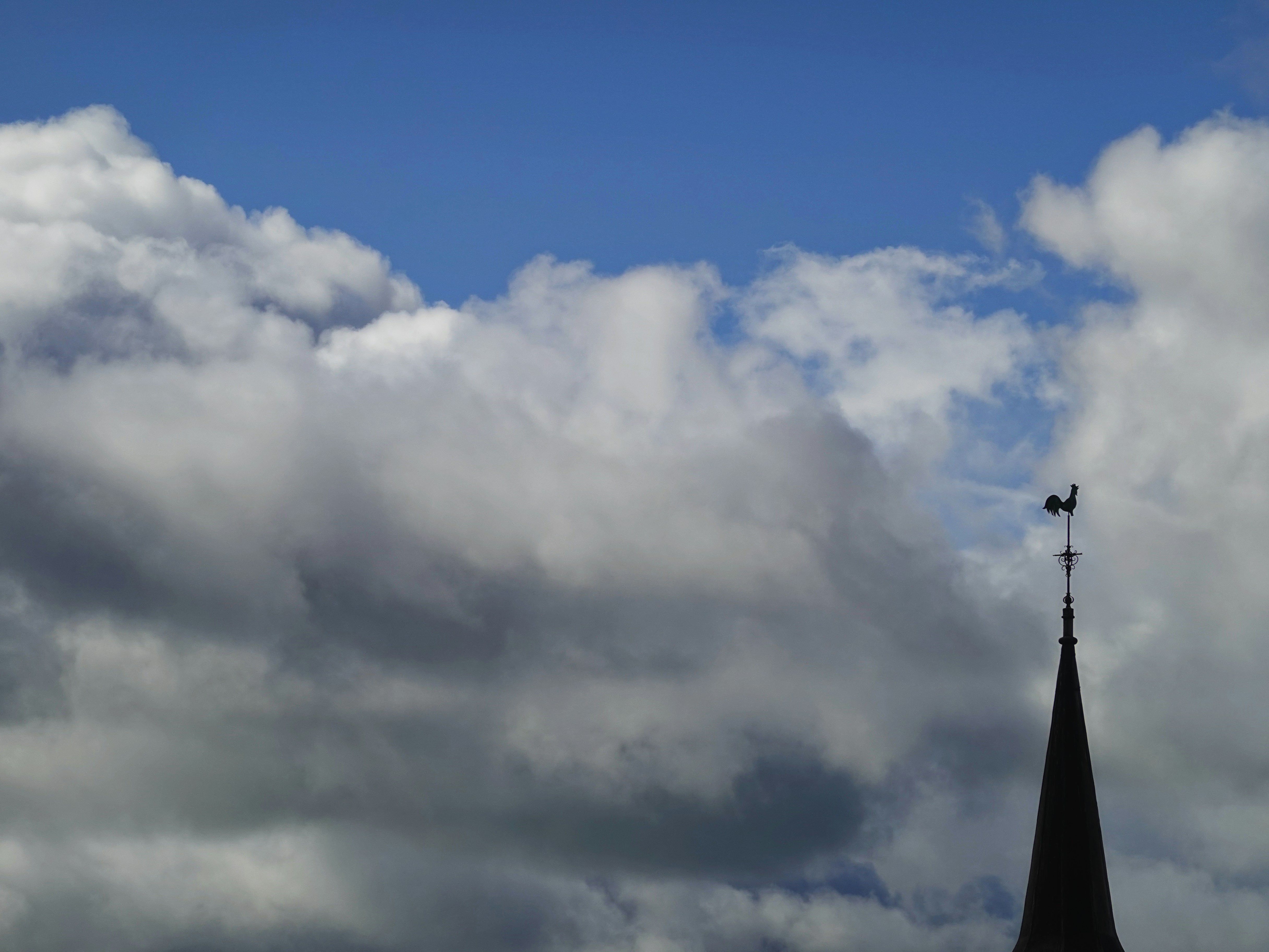 Clouds roll across a blue sky above a slender weather vane on a church spire.