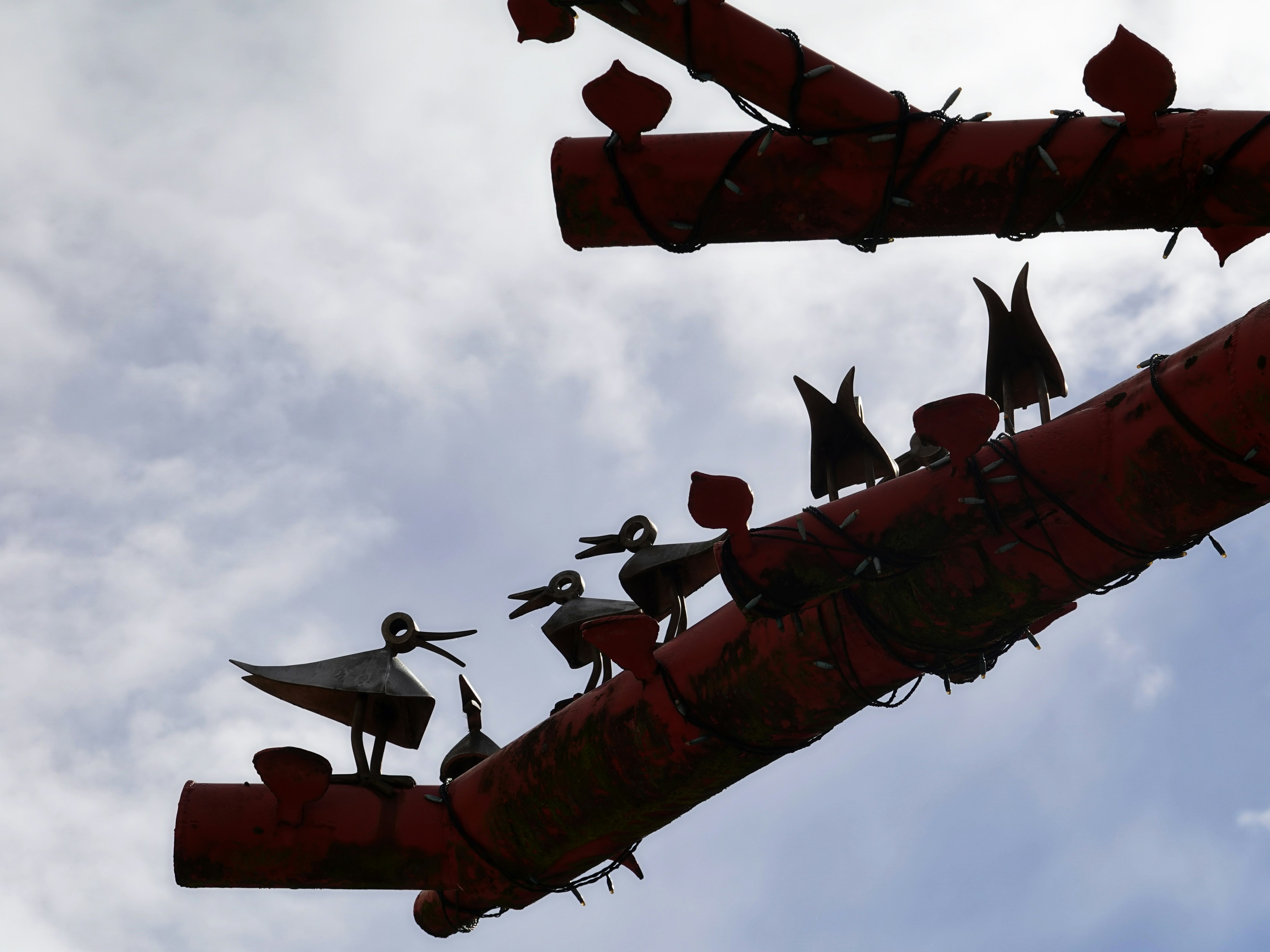 Rust-red metal sculpture forms an arching, insect-like sequence against a pale sky, with silhouetted creatures perched along the rails. The composition emphasizes linear perspective and sculptural detail.