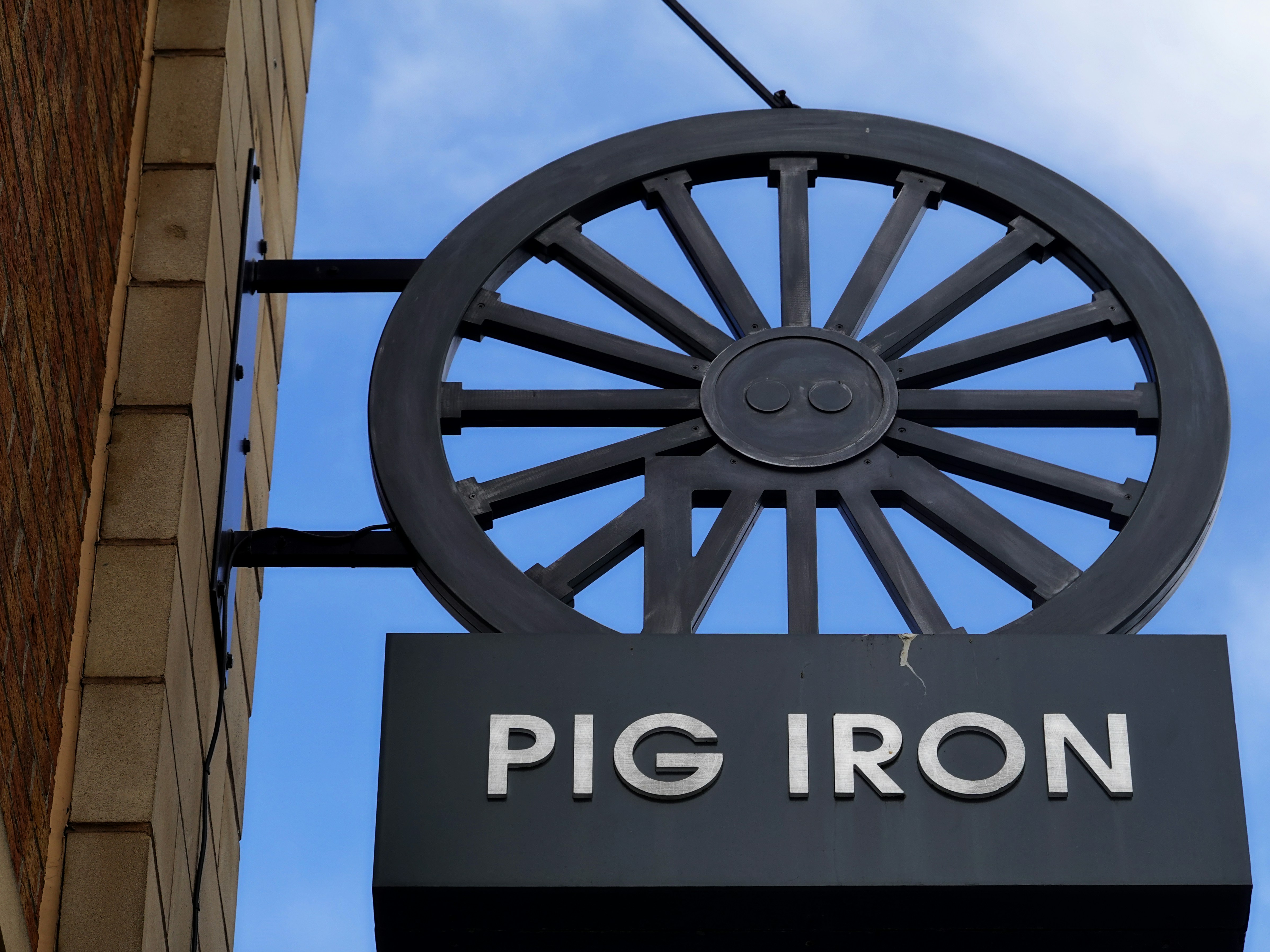 Pig Iron signboard with wheel motif against a blue sky; architectural bricks frame the composition and the metal sign sits prominently in the lower portion.