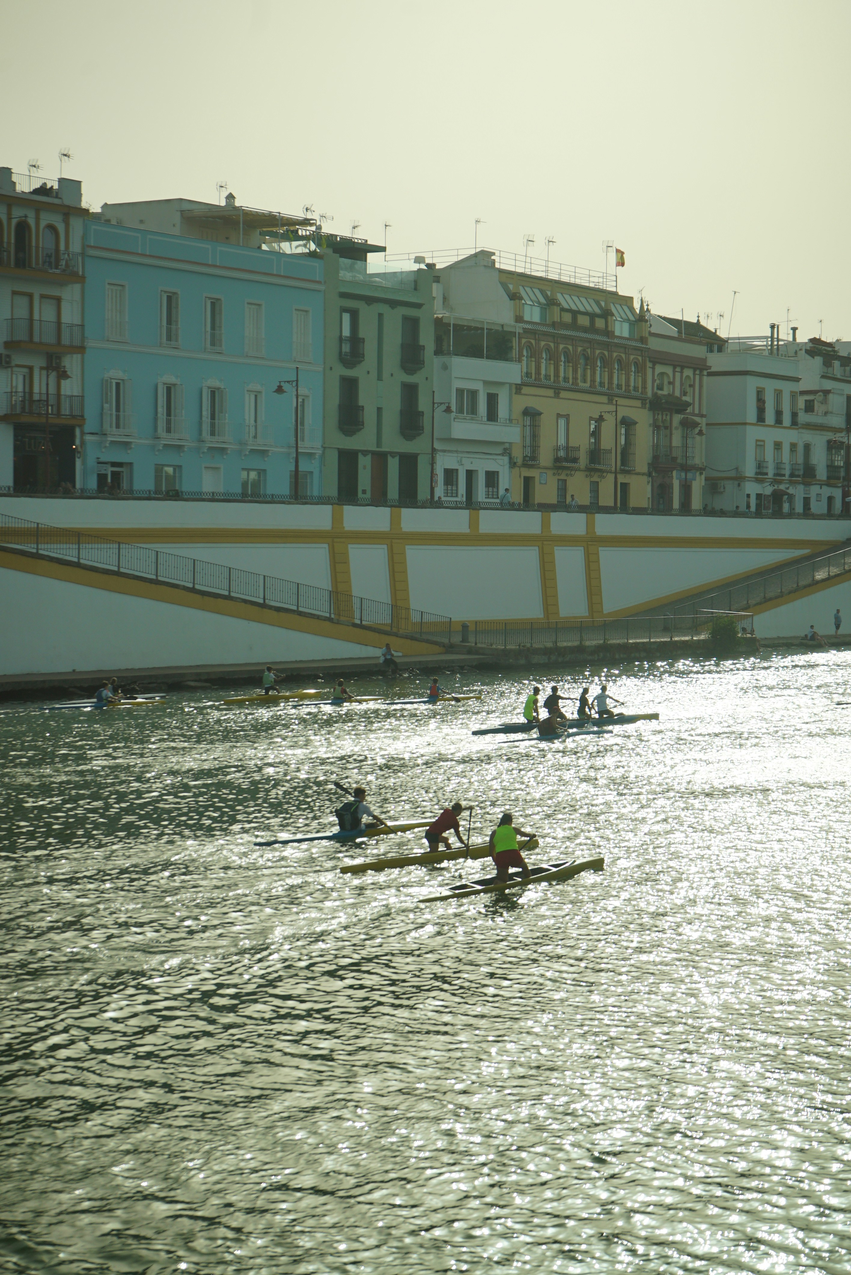 Rowers on sunlit river beneath pastel waterfront buildings