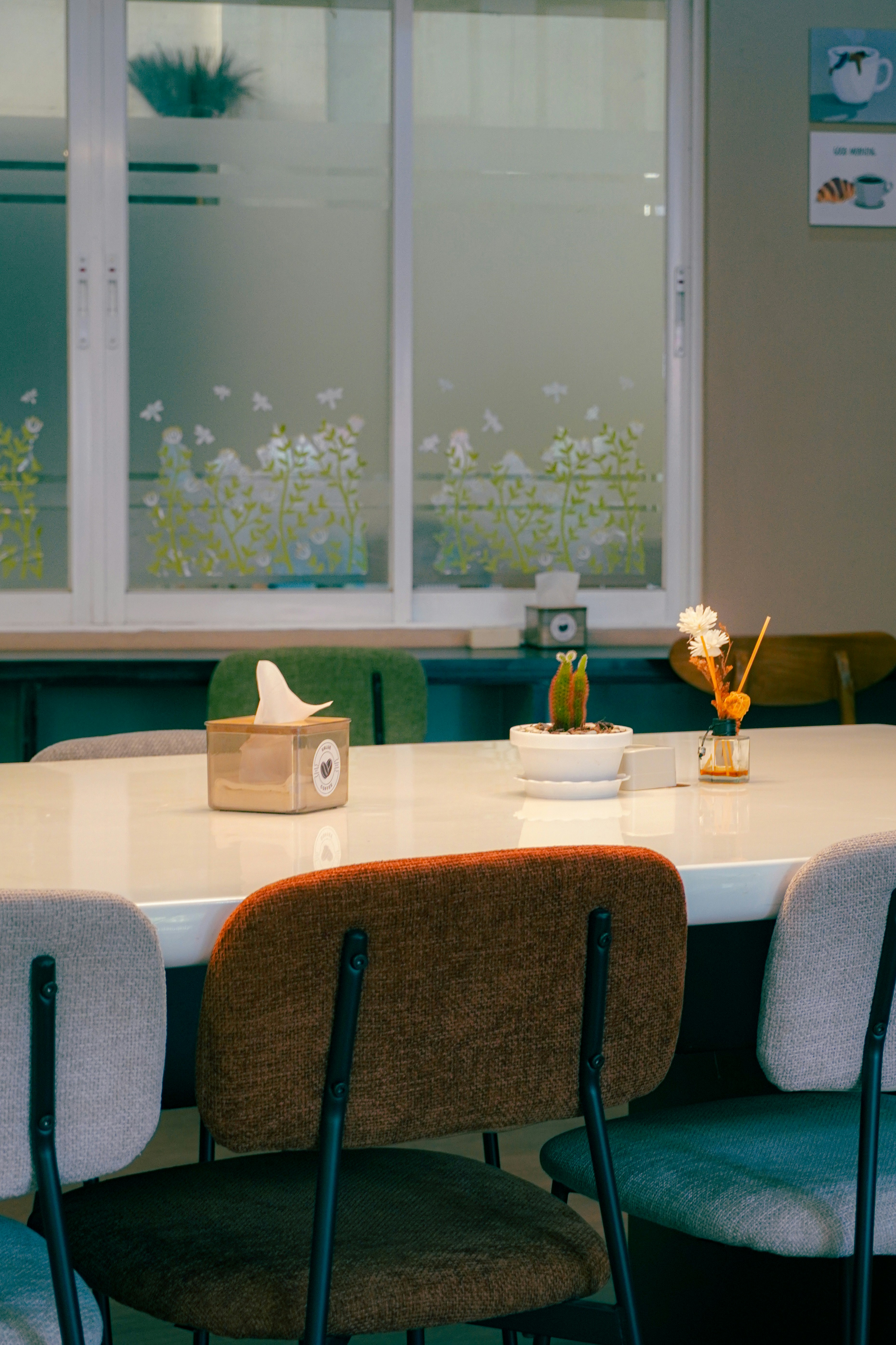 Soft-lit cafe scene featuring a white tabletop with tissue box, small potted plants, and a row of chairs; frosted windows with floral patterns in the background.