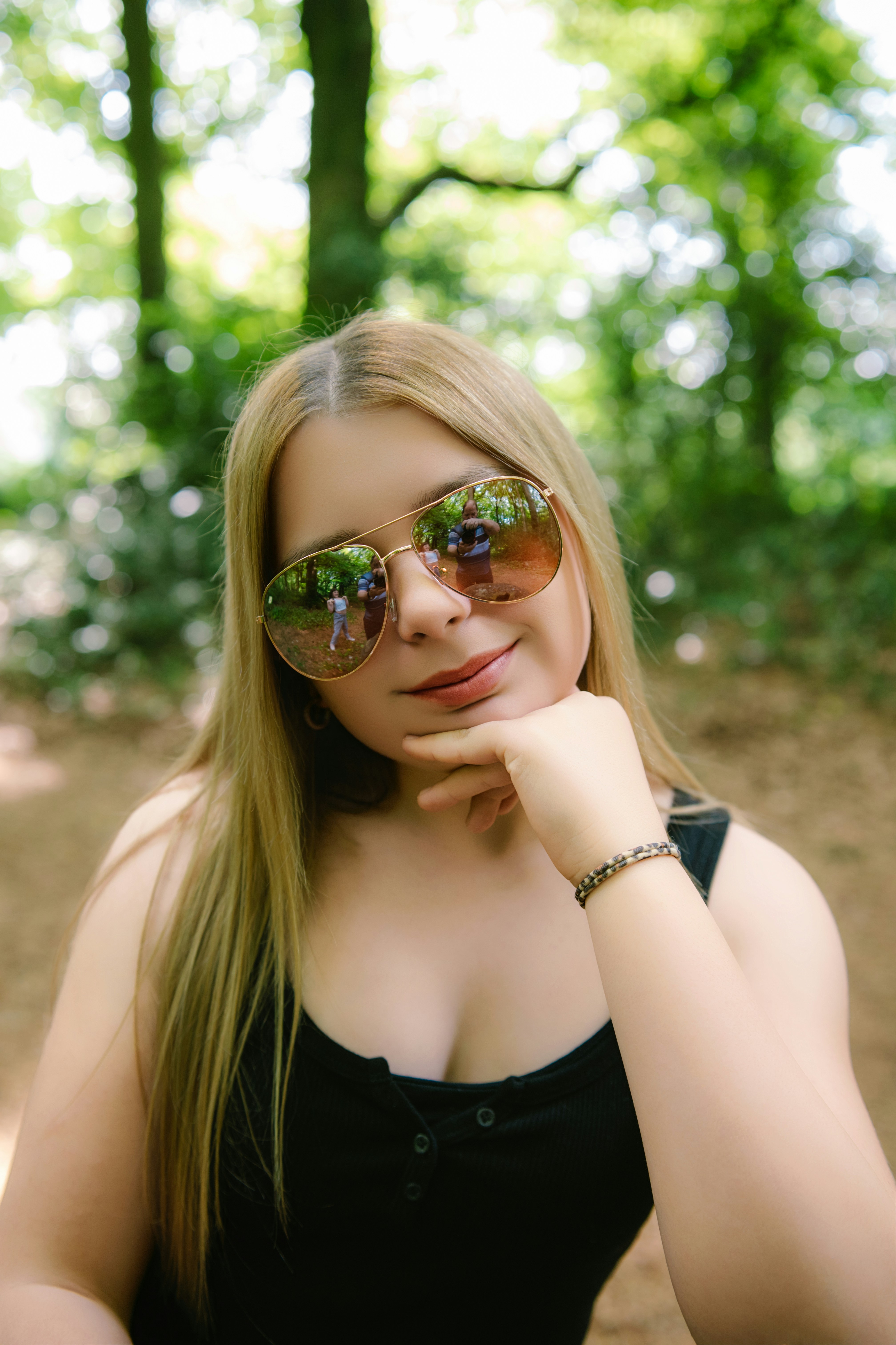 Young woman wearing aviator sunglasses sits in a sunlit forest, head resting on her hand, black sleeveless top, soft bokeh background.