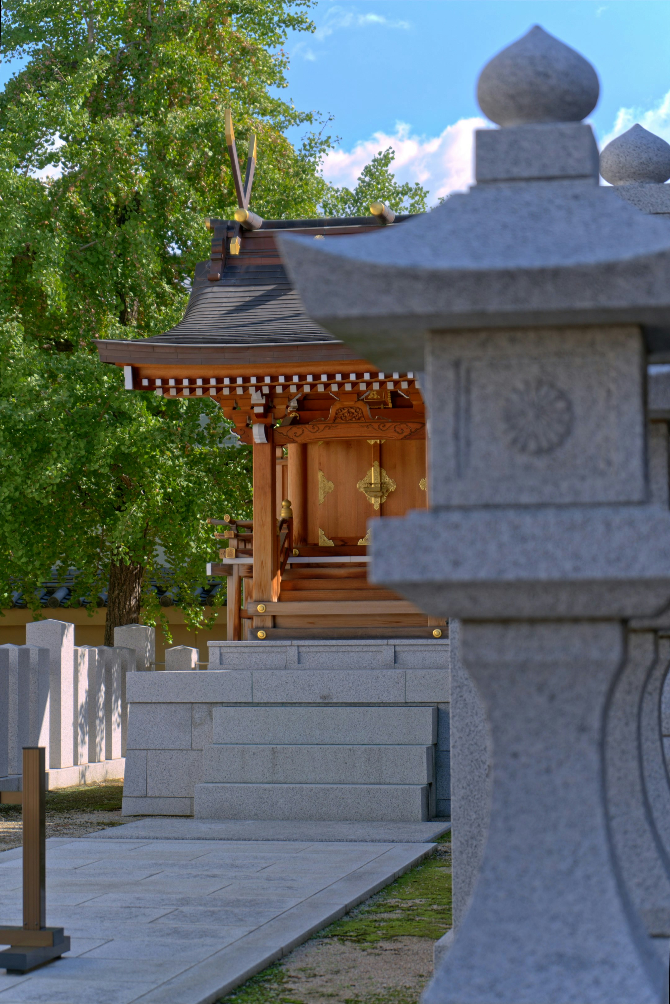 Stone lanterns frame a distant wooden shrine with ornate detailing and a sloped roof, peeking through lush green trees.