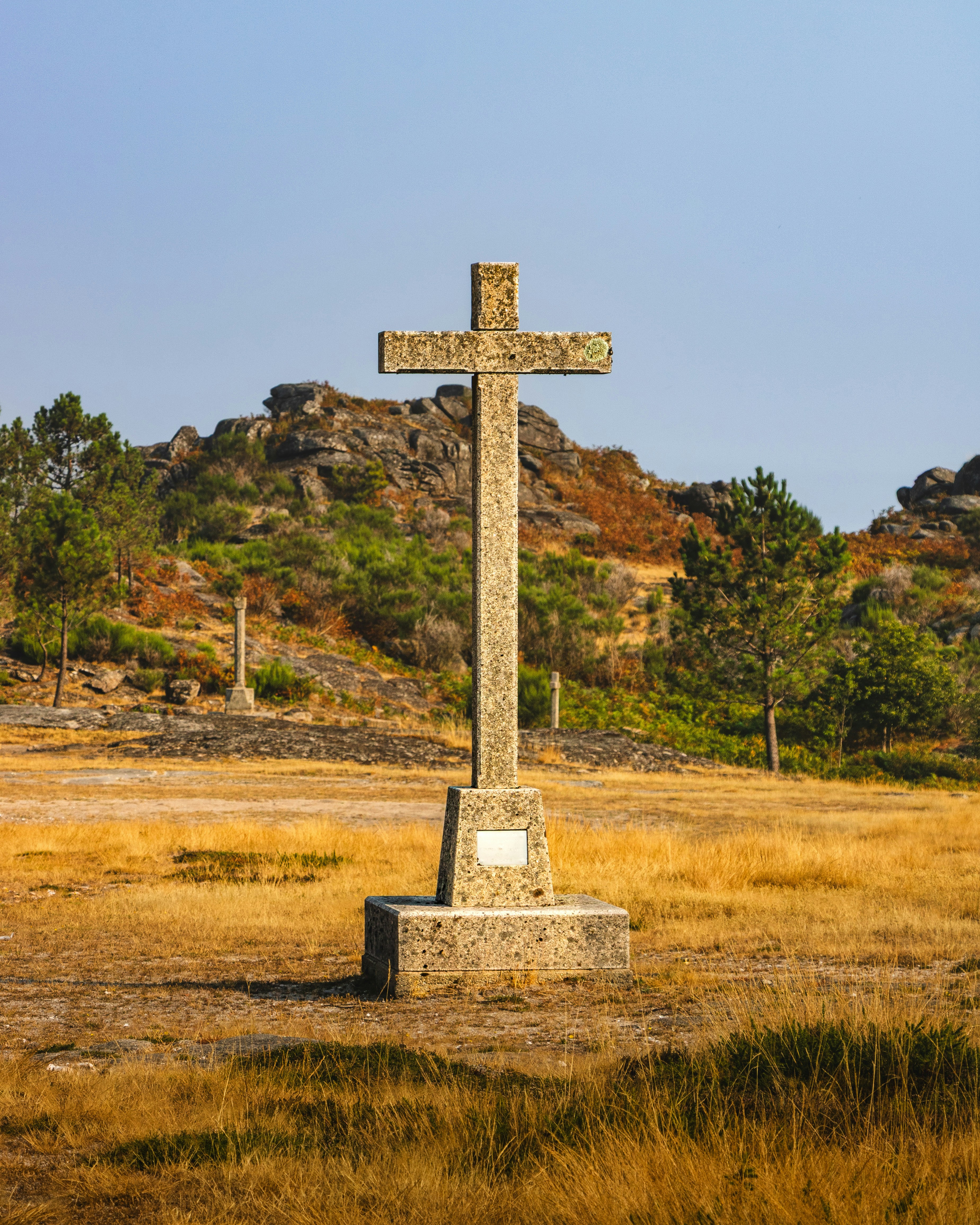 Weathered stone cross standing in a sunlit field with sparse grass and distant rocky hills; a quiet landscape that may be part of a park or memorial site.