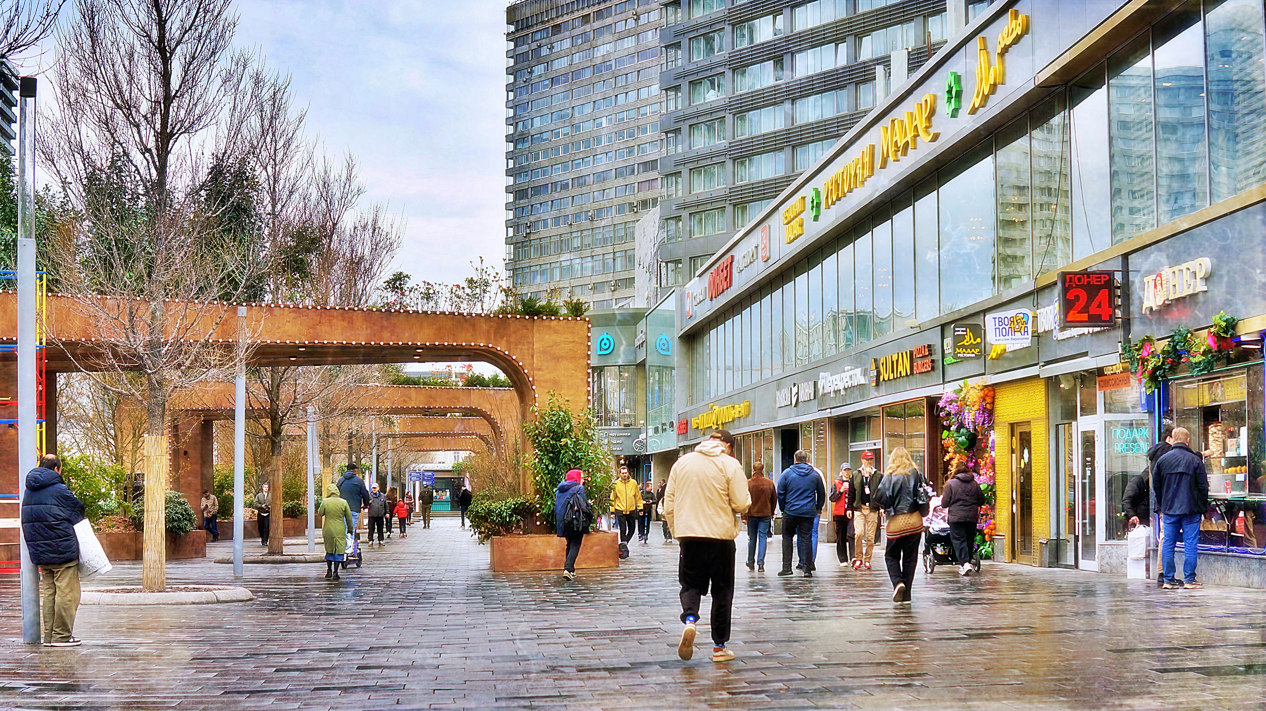 Busy pedestrian street with storefronts and glass facades on a wet day; pedestrians walk under shaded arches along a modern shopping area.