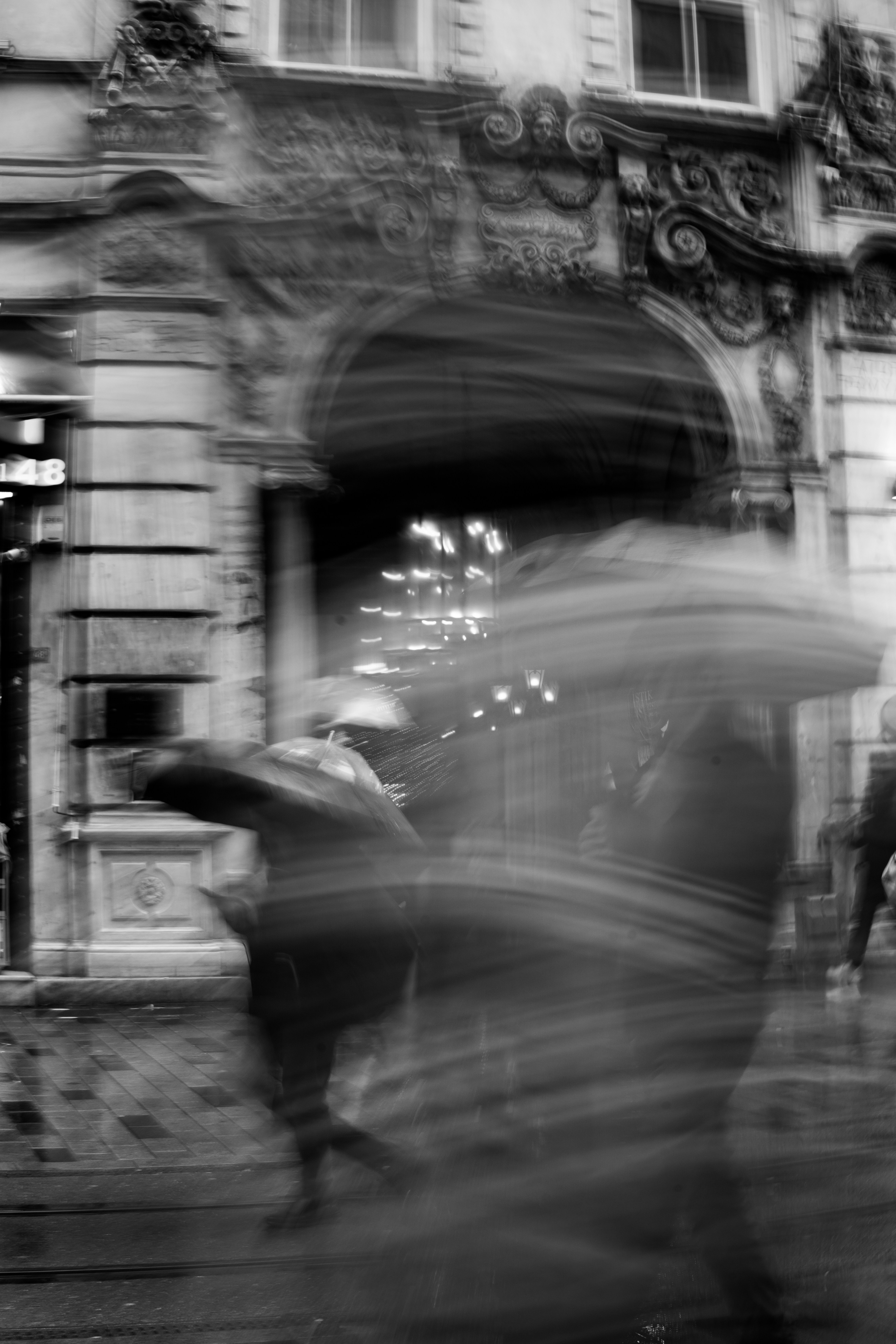 Black-and-white street scene with blurred figures moving past an ornate building entrance, conveying motion and urban energy.