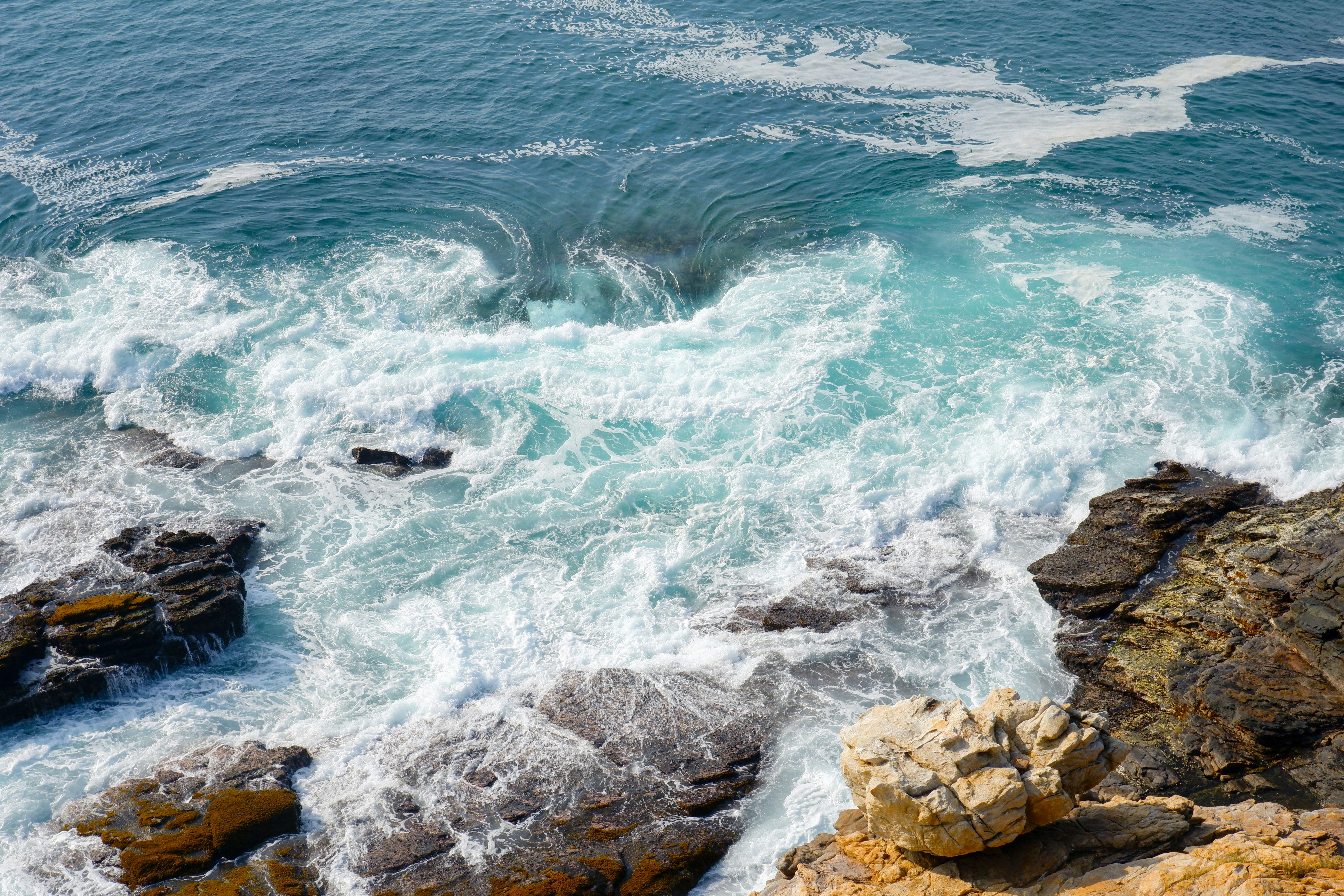 Crashing surf against jagged rocks along a sunlit coastline, highlighting turquoise water and foamy white spray.