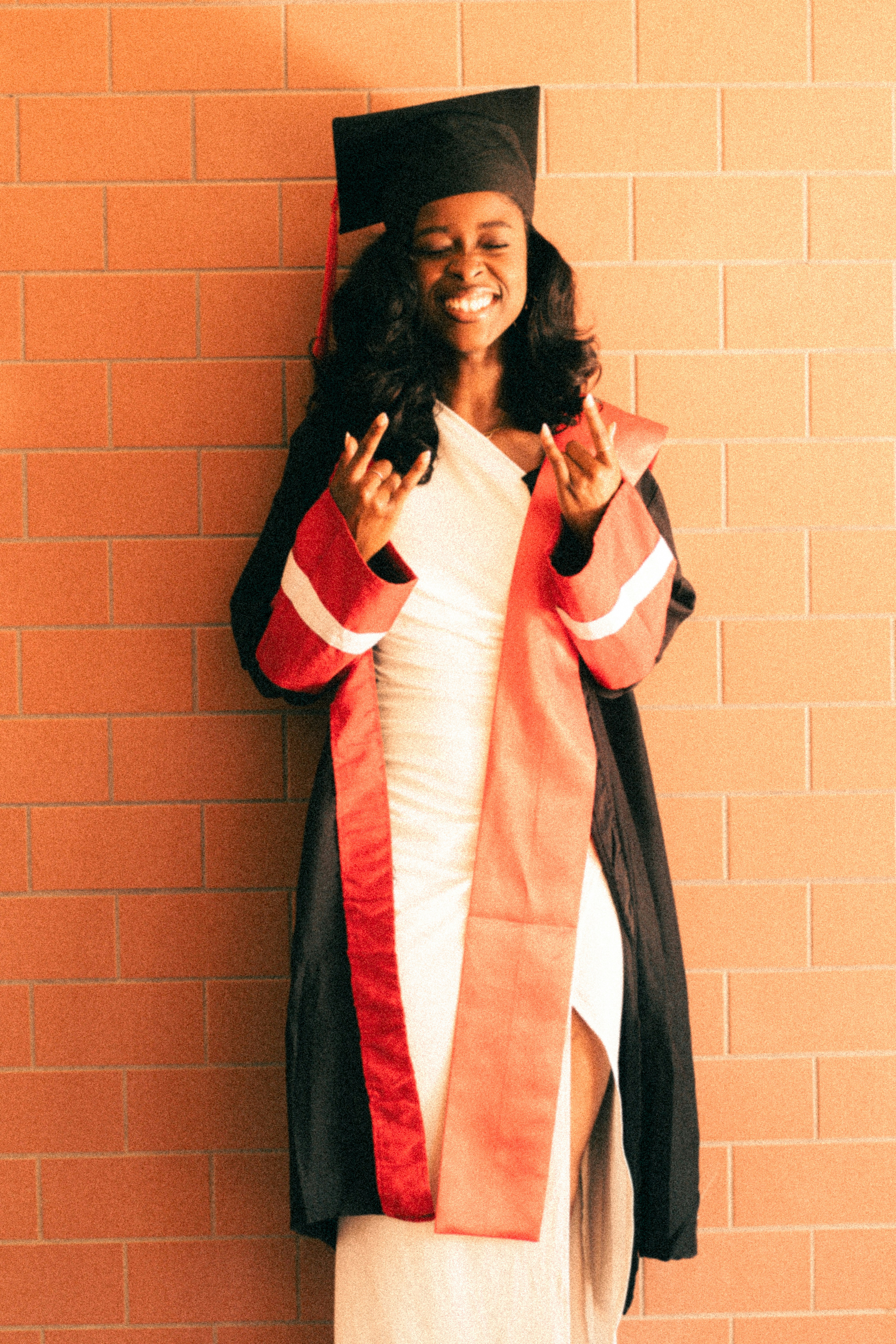 Joyful graduate in a sunlit brick hall, flashing a double peace sign