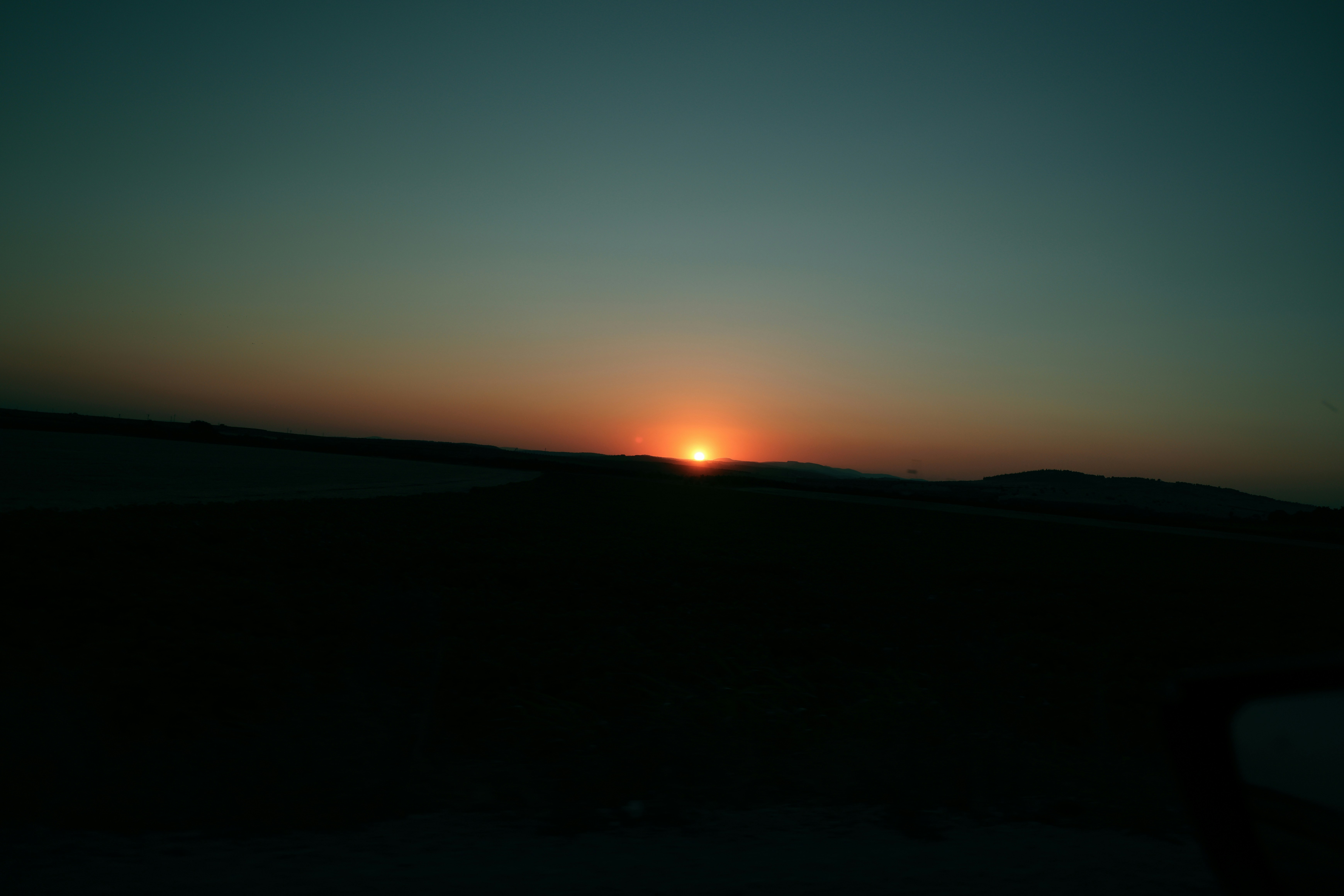 A low-contrast desert horizon at sunset with a bright orange sun just above the dark silhouetted dunes; the scene emphasizes the expansive sky and distant landforms. This is a natural landscape capture.