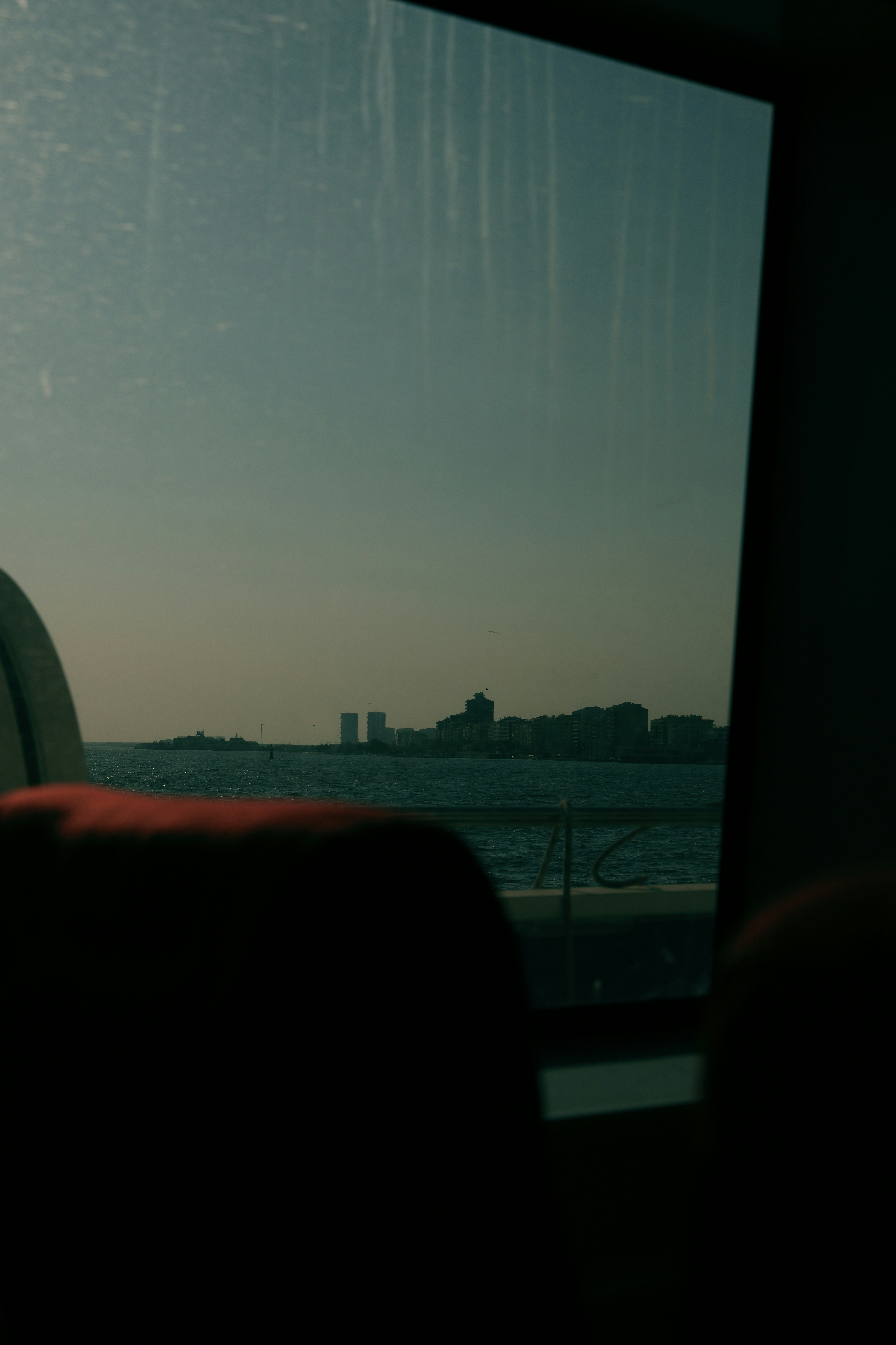 Softly lit sea view framed by a window, with distant city skyline and foreground silhouettes; photograph captures the calm water and hazy horizon from an interior vantage point.