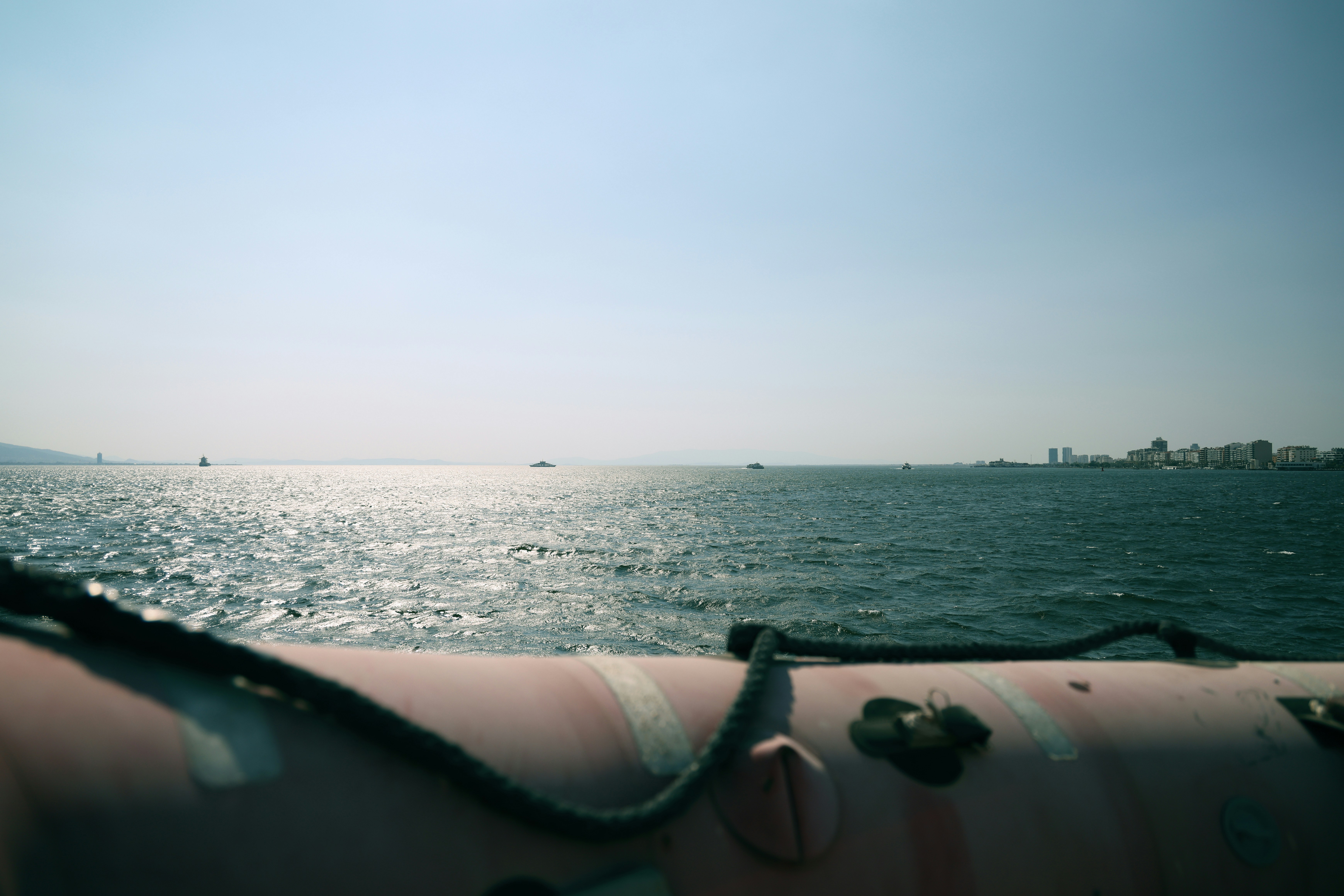A wide seascape seen from a boat deck, with the horizon and glistening water occupying the upper two-thirds and a blurred lifebuoy and rope in the foreground. A distant shoreline and small boats punctuate the horizon.