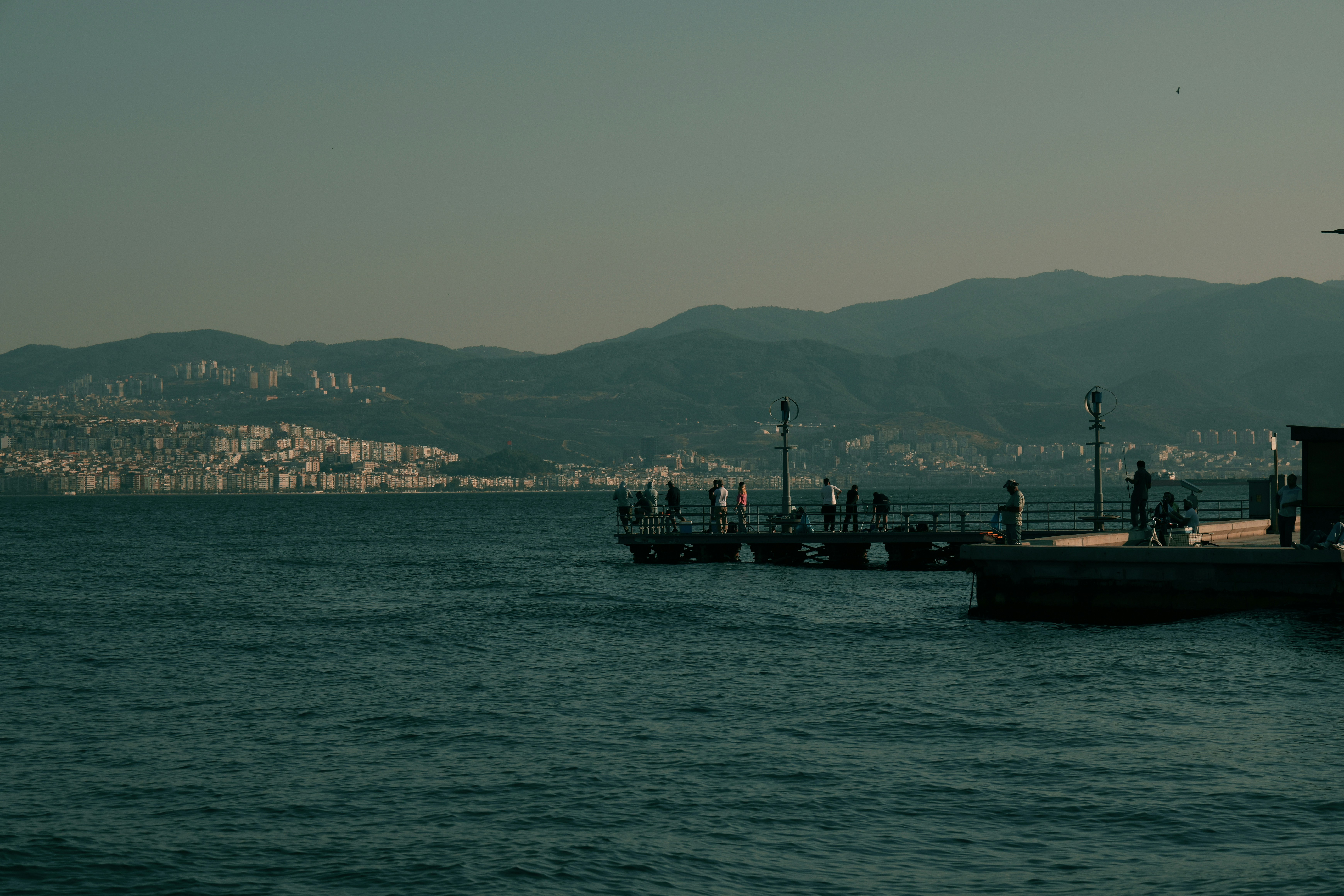 Harbor with piers and distant cityscape along a calm coastline at dusk; a few figures on the pier and a ship silhouette against a mountain backdrop.