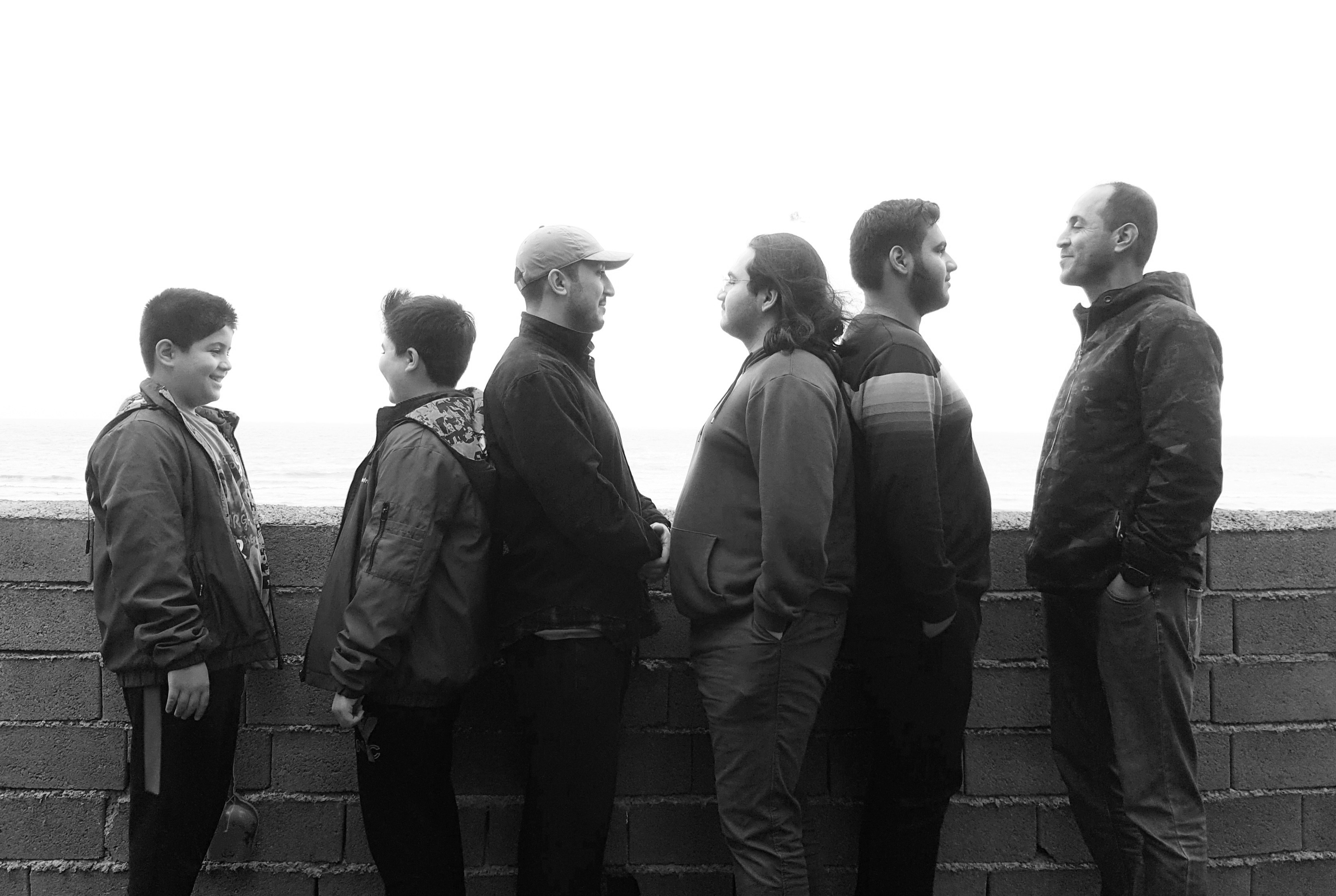 Seven individuals stand in profile along a brick barrier, forming a linear silhouette against a pale horizon; the scene emphasizes unity and variation in stance. This is a monochrome group portrait.