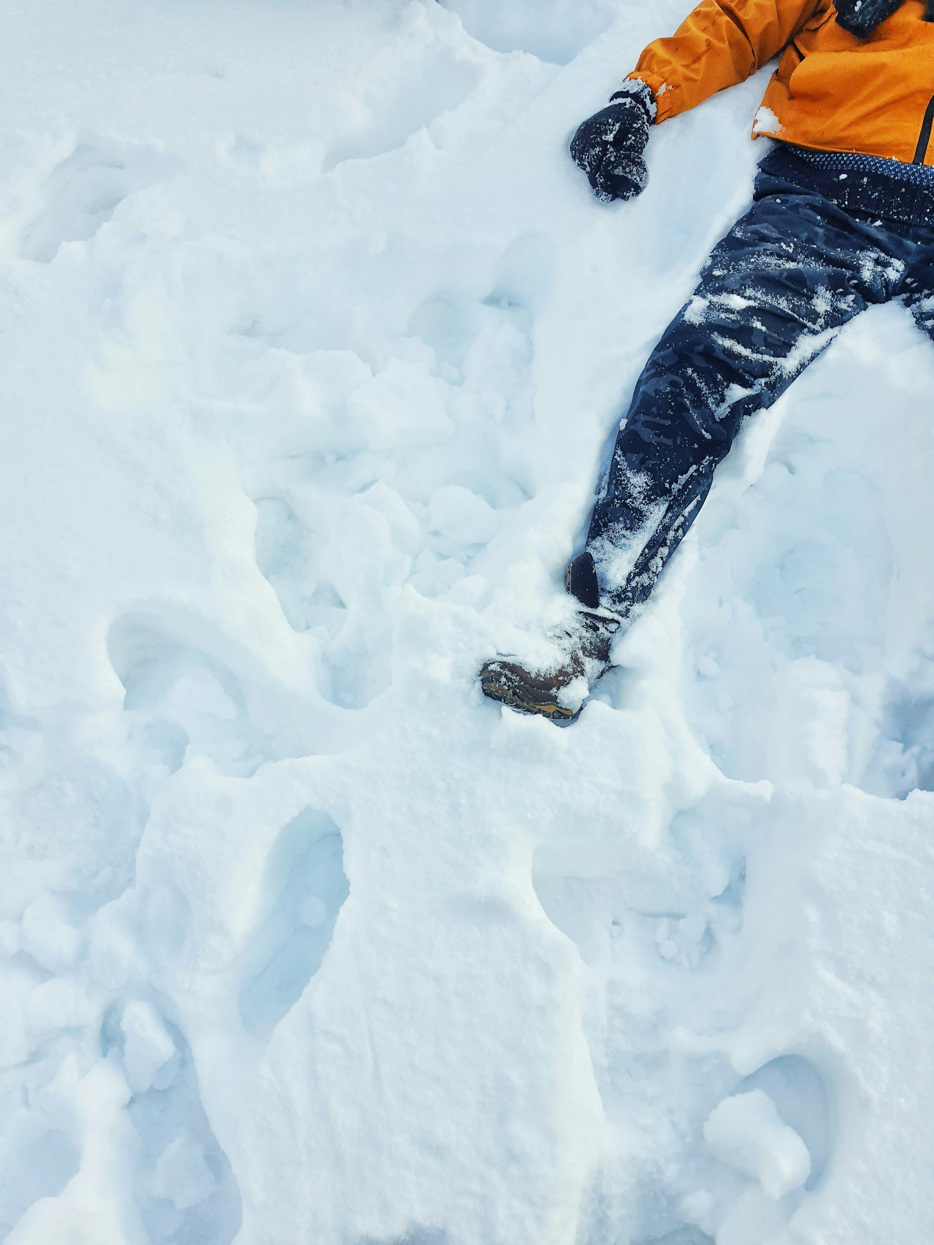 Partial figure in orange jacket lies in deep snow, booted leg and gloved hand visible among fresh drifts.