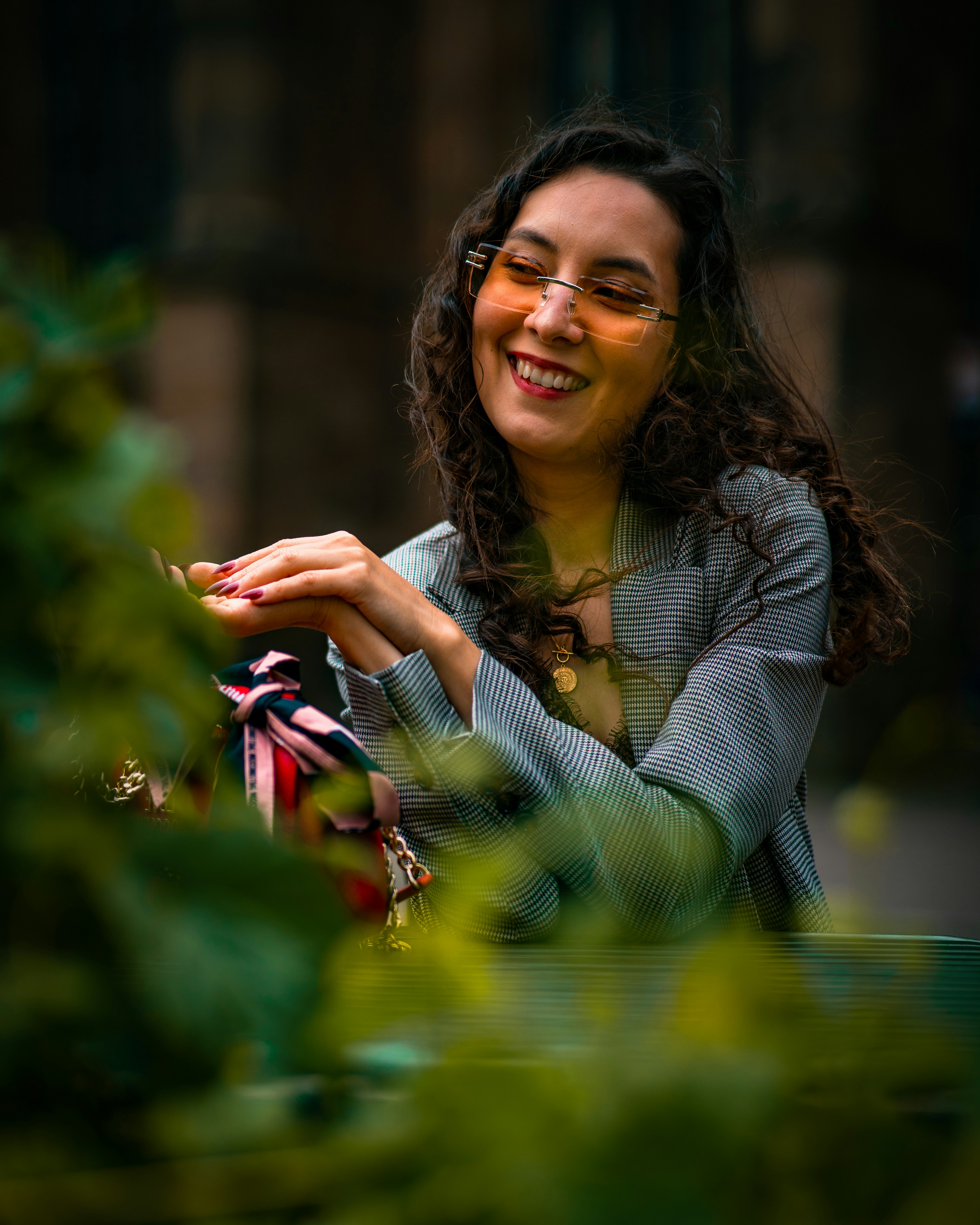 Smiling woman with glasses sits outdoors, hands resting on a colorful bag, with shallow depth of field and warm tones in a garden setting.