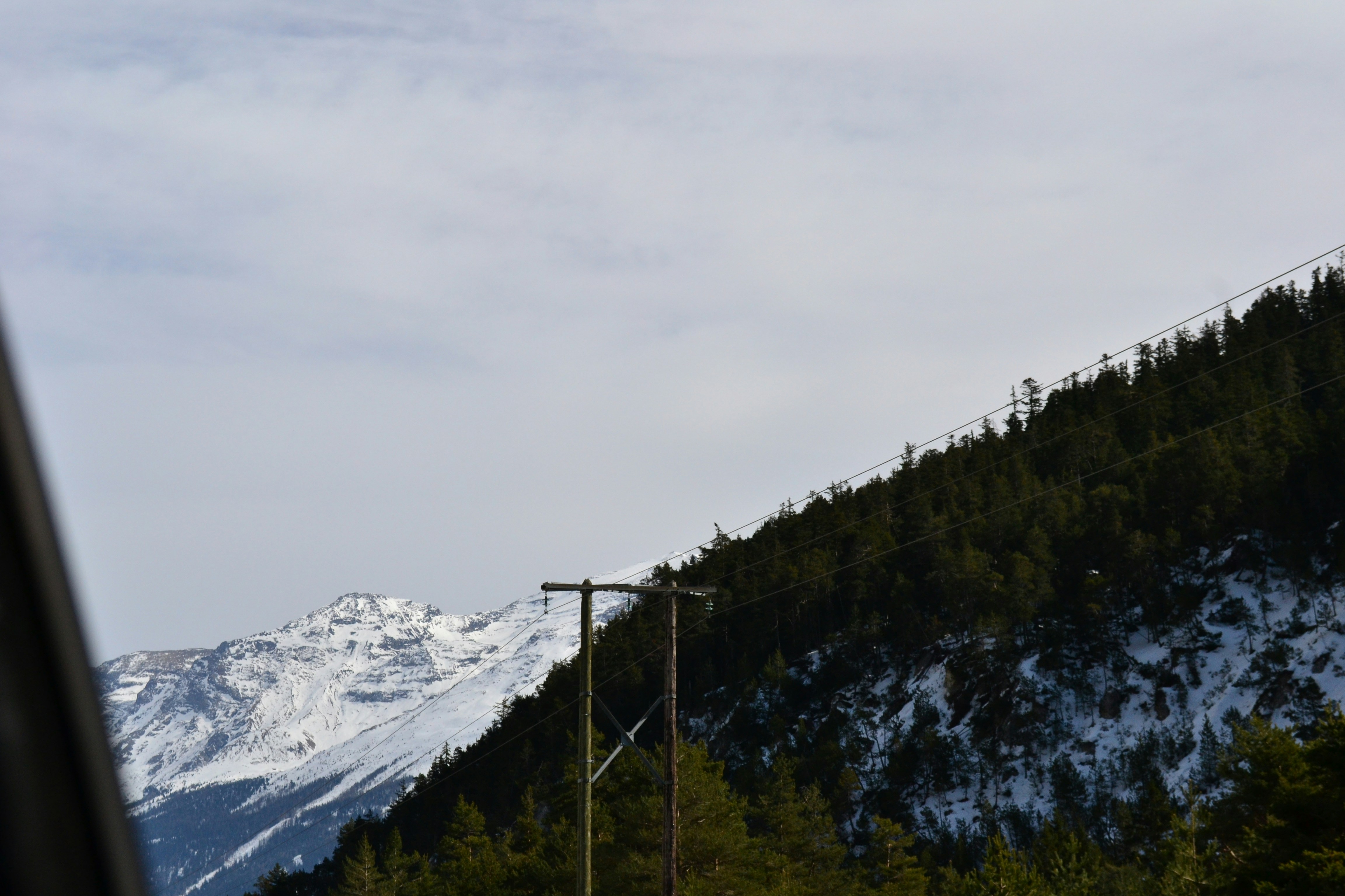 Snow-dusted mountains peek through a car window, with a power pole and hillside in the foreground, captured on a bright day.