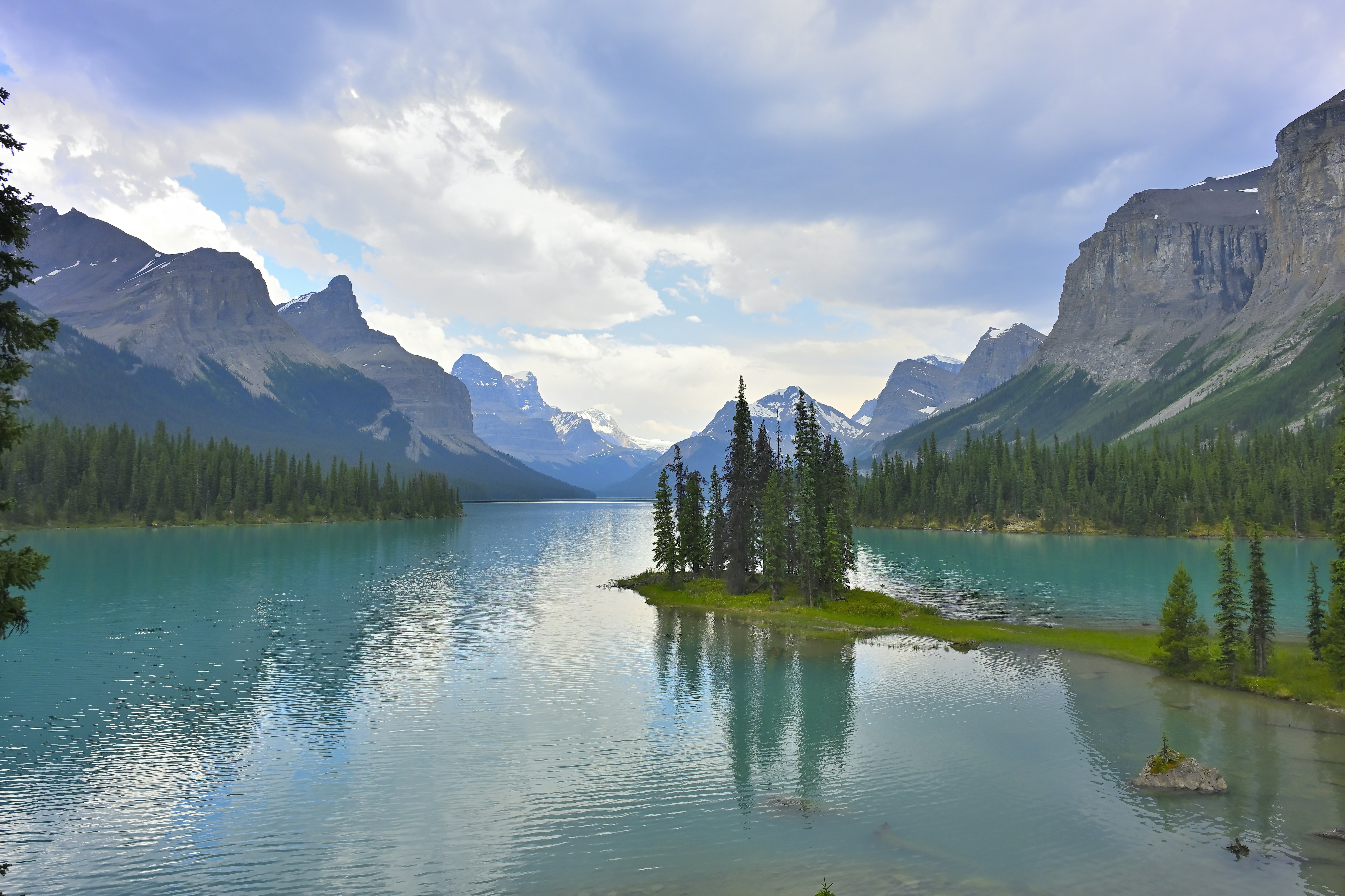 Clear turquoise lake with a small pine island set against a backdrop of jagged mountains under a bright sky; a serene landscape scene highlighting water, forest, and alpine peaks.