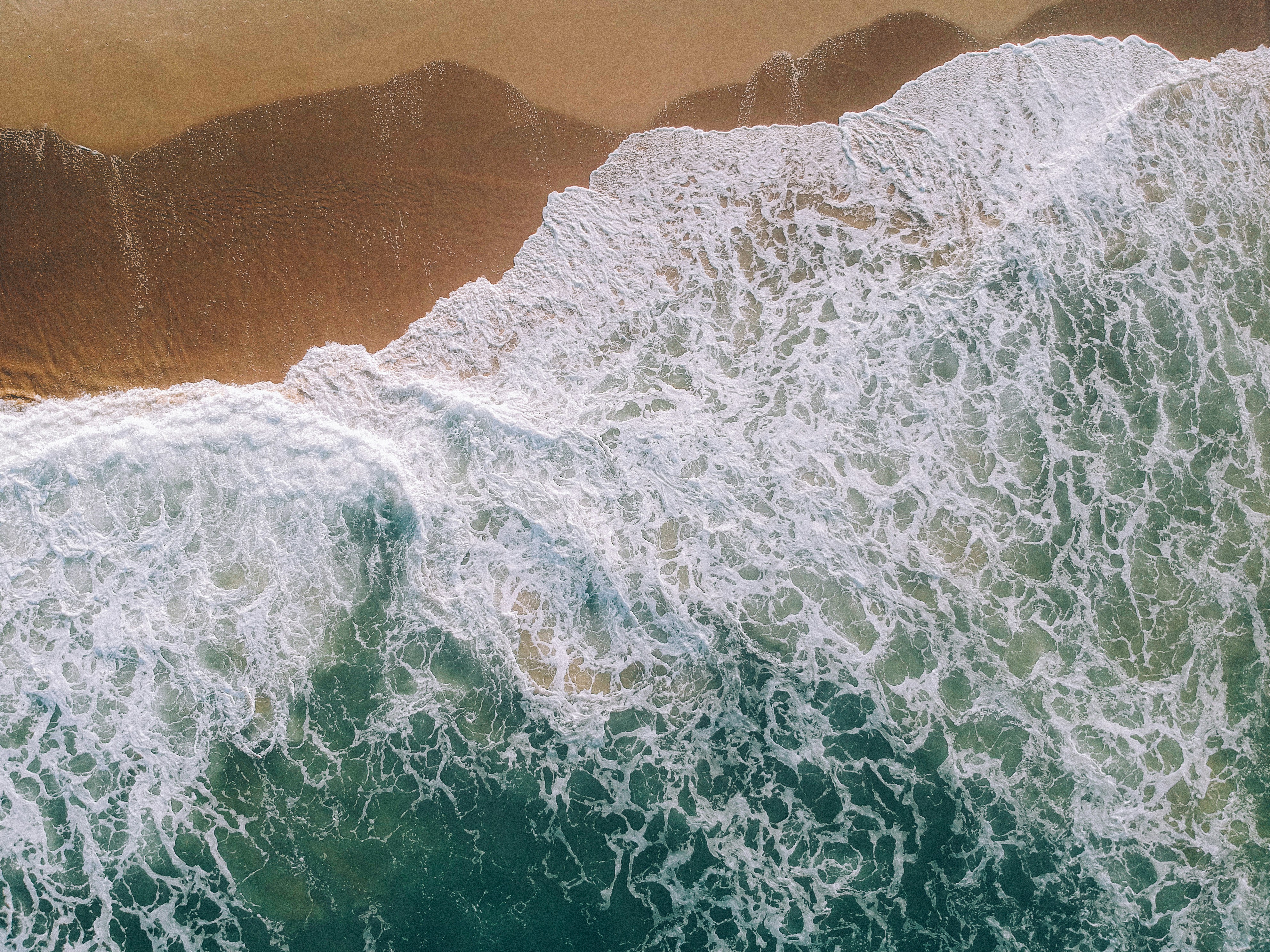 Crashing white-tipped waves sweep onto a sandy shoreline, capturing the dynamic boundary between sea and beach in bright daylight. Aerial perspective emphasizes the texture of foam and turquoise water.
