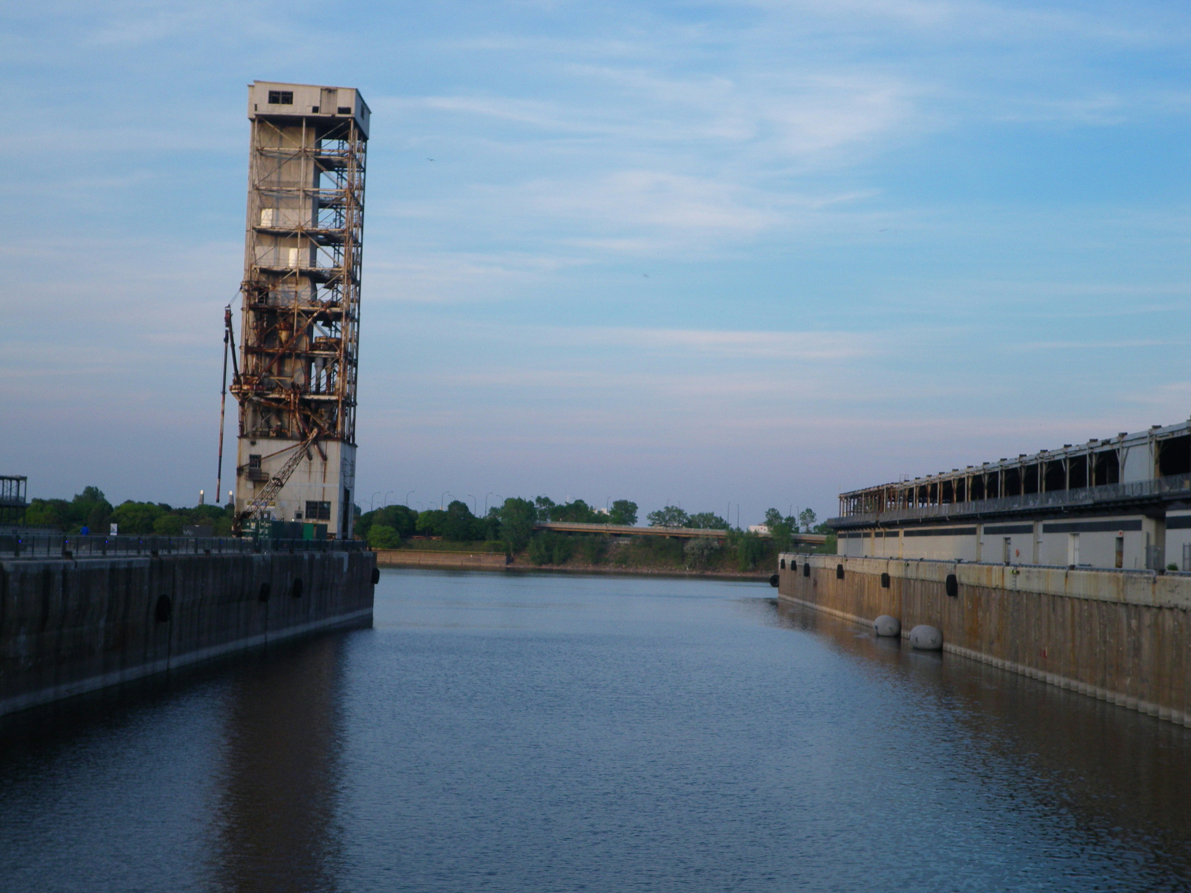 Industrial ruin beside a calm waterway with a tall, skeletal structure on the left and a long, weathered quay on the right, captured in soft daylight.