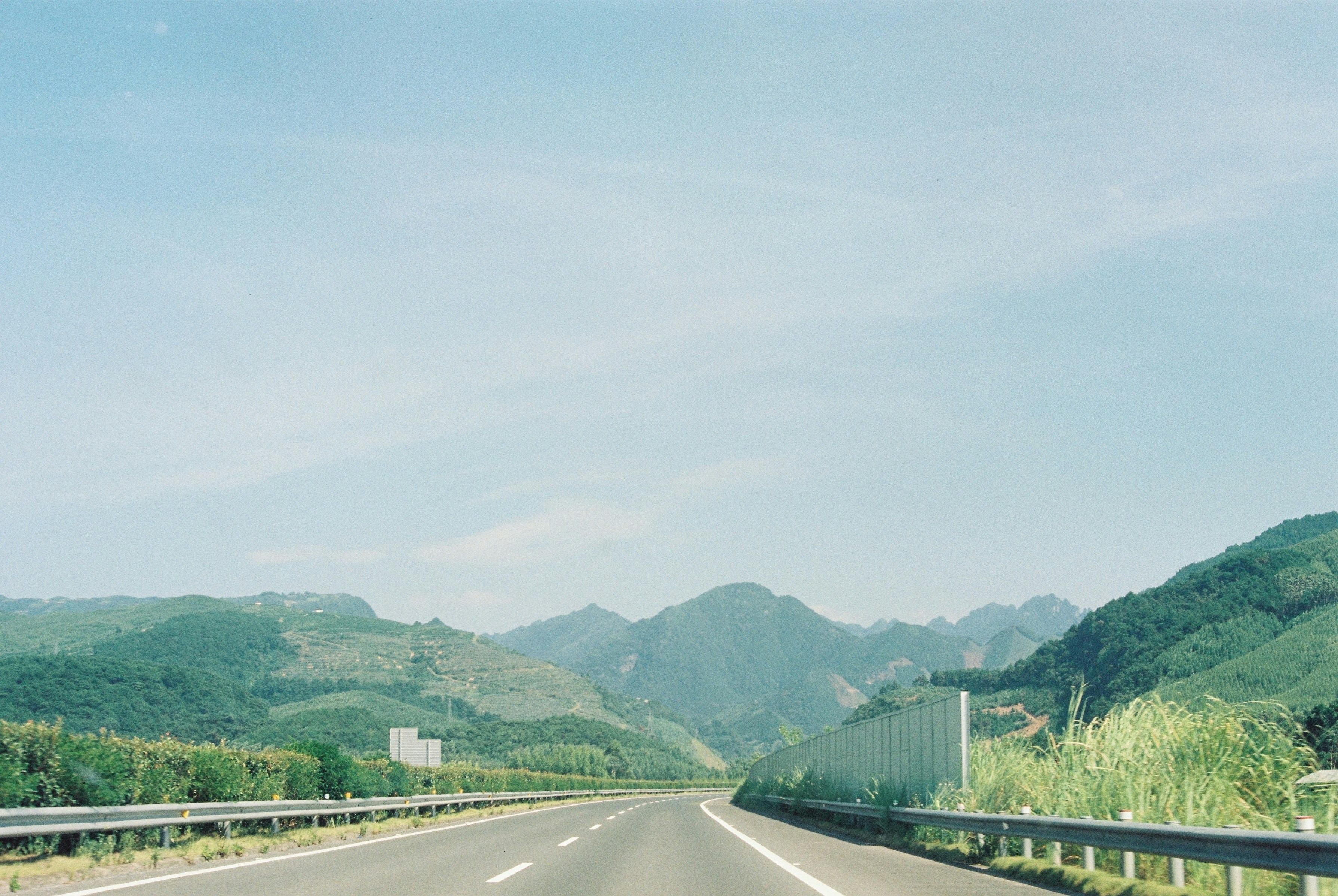 Wide highway leading toward distant mountains under a clear blue sky, framed by guardrails and green countryside.