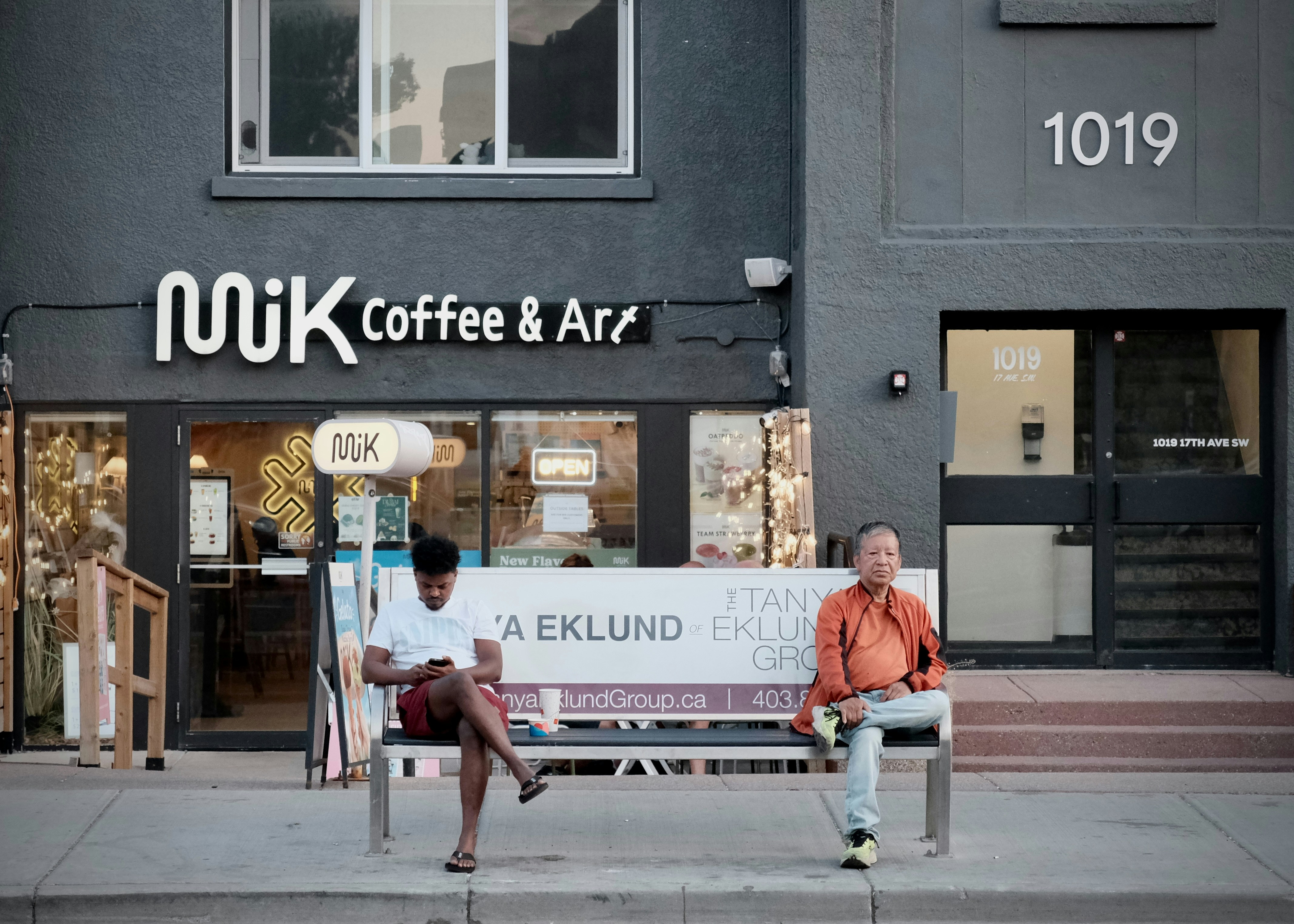 Two strangers on a sunlit bench outside a coffee shop with a bold storefront sign
