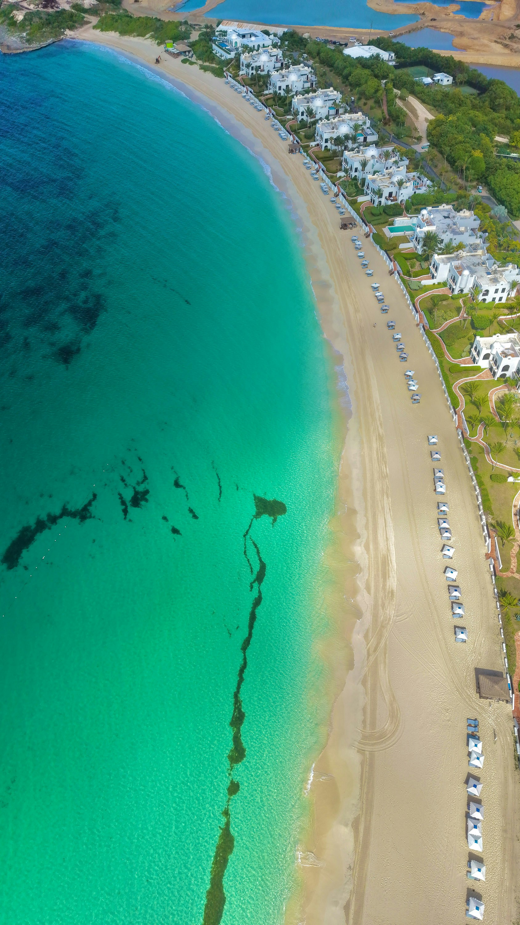 Aerial view of a turquoise sea meeting a sandy beach lined with white resort villas and loungers. A curved shoreline and palm-lined promenade define the coastal resort scene.