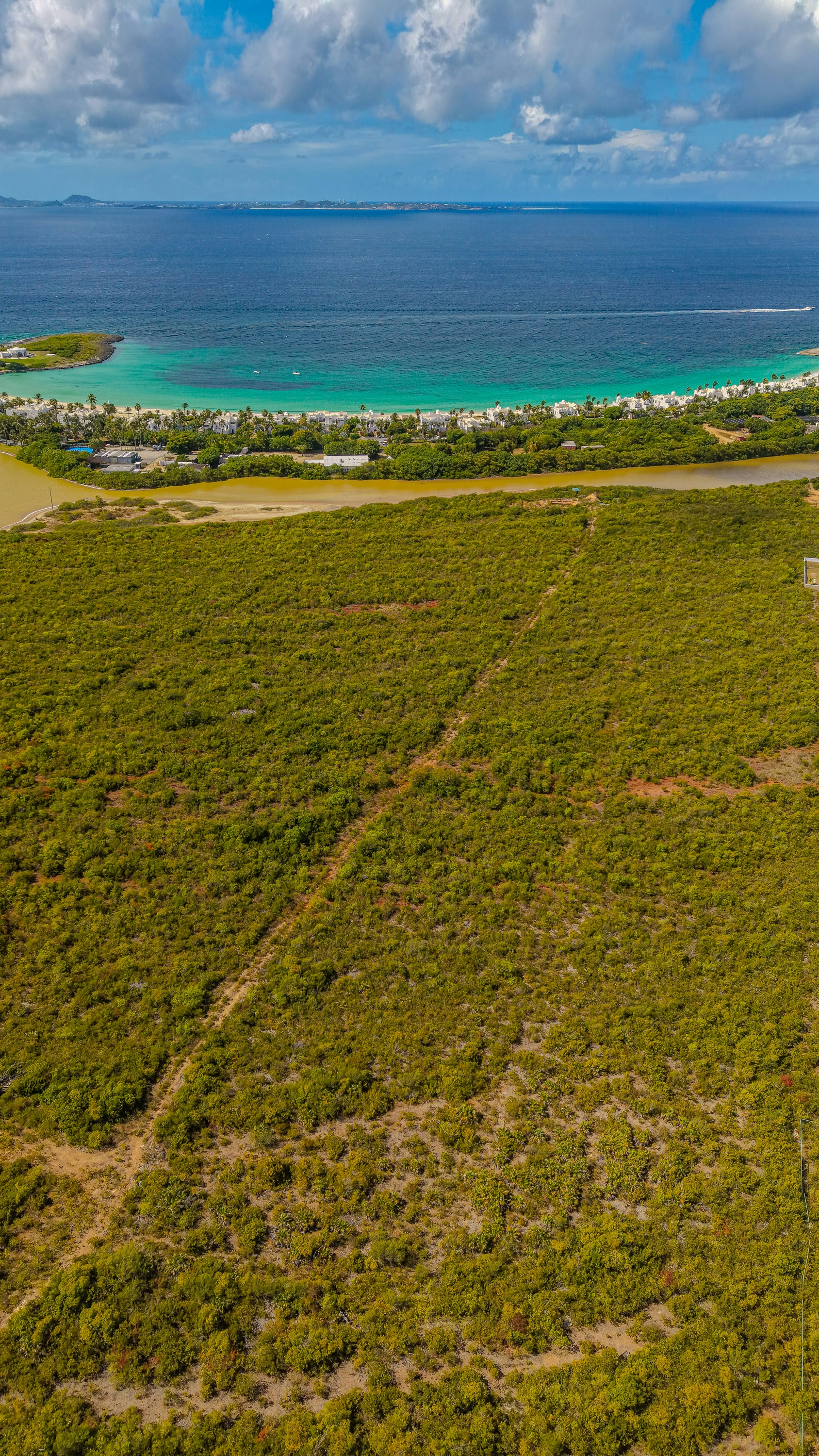 Aerial view over turquoise coastal shallows meeting a green scrubland, with a distant urban fringe along the shore; the scene captures a broad expanse of beach, sea, and undeveloped land.