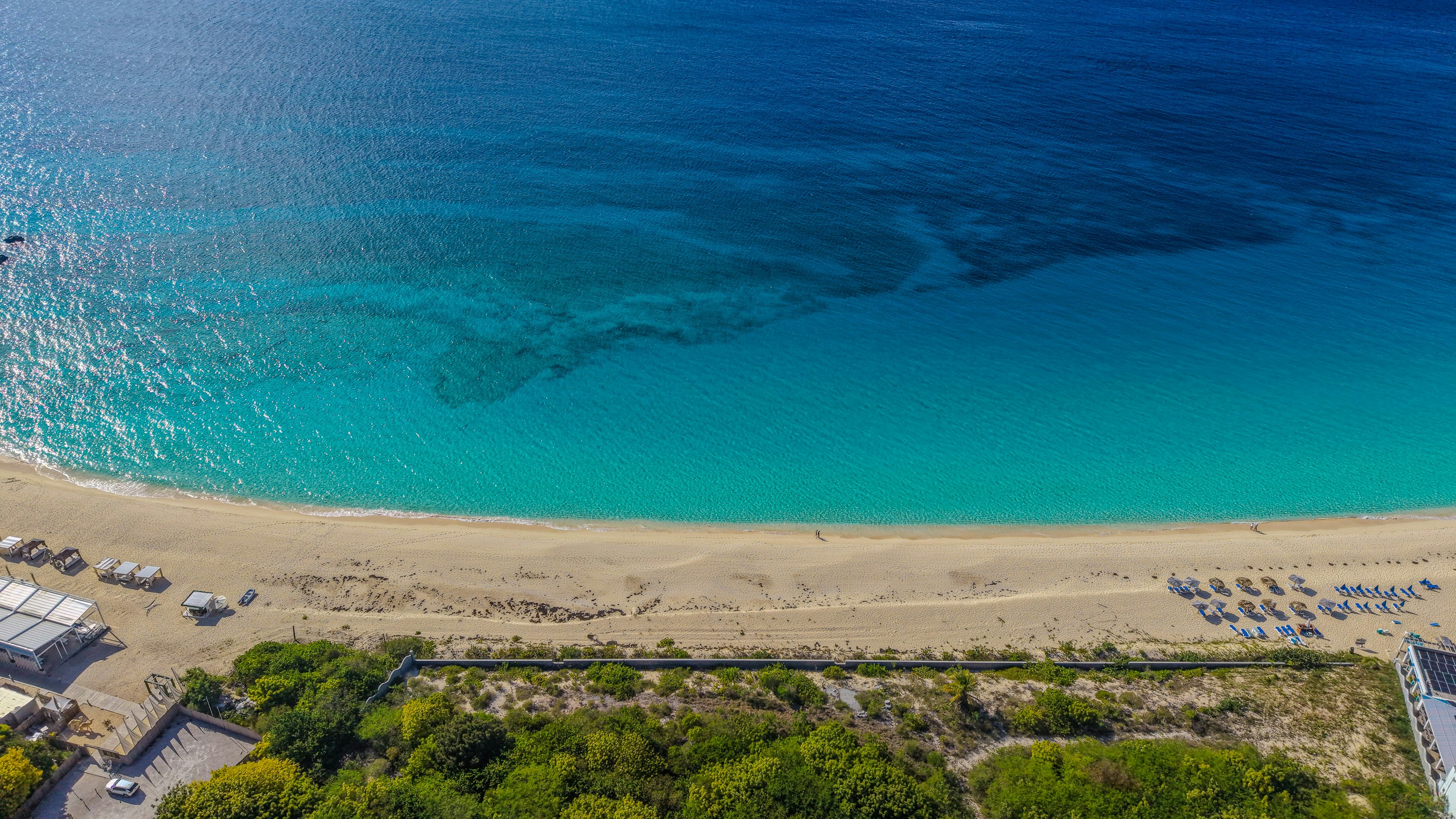 Aerial view of a turquoise sea meeting a sandy beach with rows of umbrellas and a green coastal strip, illustrating a tranquil resort scene.