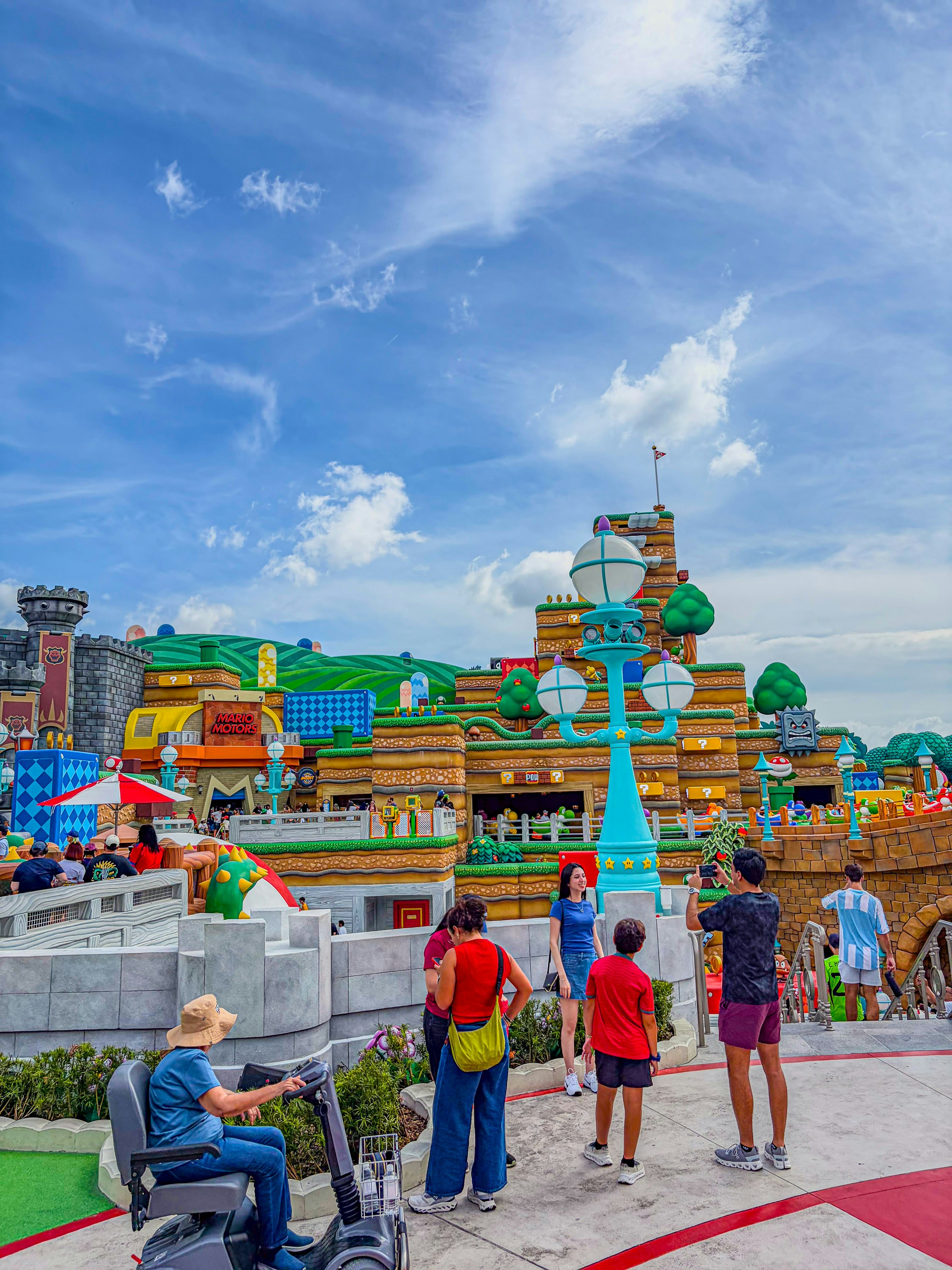Vivid theme-park facade with whimsical castle-like structures and visitors foreground; a motorized scooter and groups of people emphasize scale and activity.