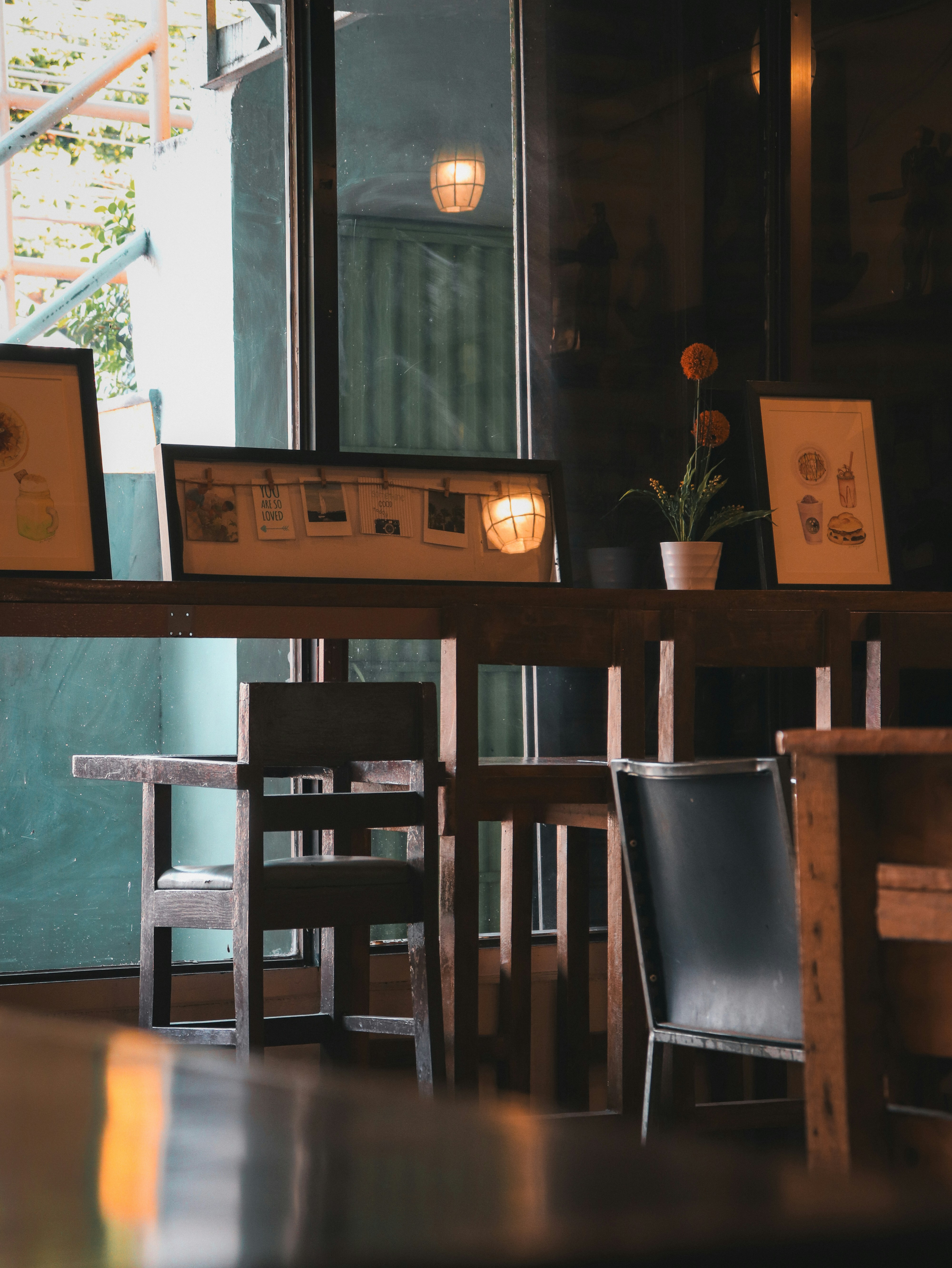 Muted cafe interior with wooden tables and a single potted plant under warm pendant light, framed by windows and art on the shelf.