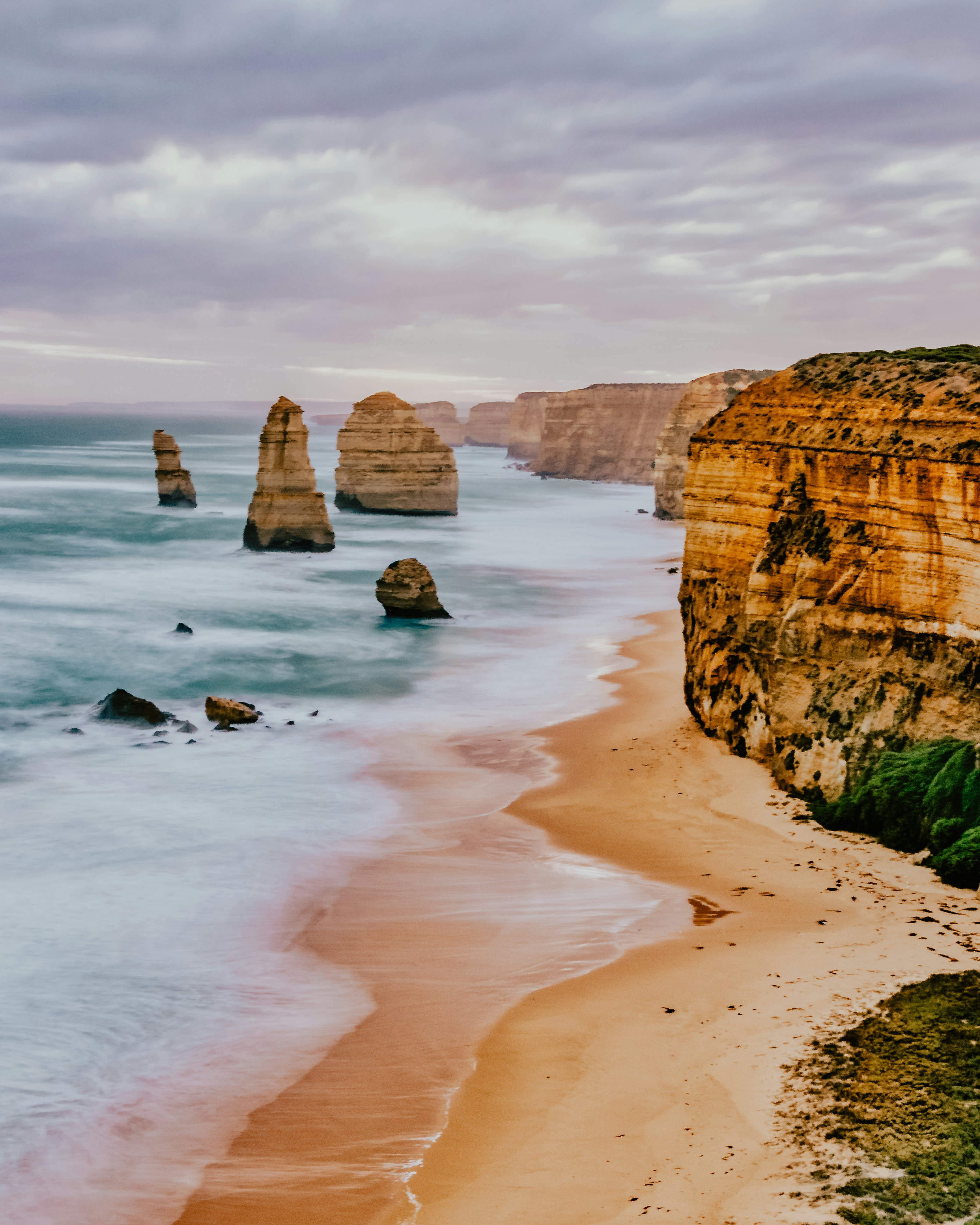 Elevated coastal view of the Twelve Apostles on Victoria's Great Ocean Road, Australia. Limestone sea stacks rise from milky turquoise water in the left half of the frame, while a tall ochre cliff face dominates the right, streaked with iron oxide and topped with green scrub. A long exposure smooths the incoming surf into soft white bands curving along the empty apricot sand. Overhead, layered grey and lilac cloud at dusk. Muted, cinematic colour grade.