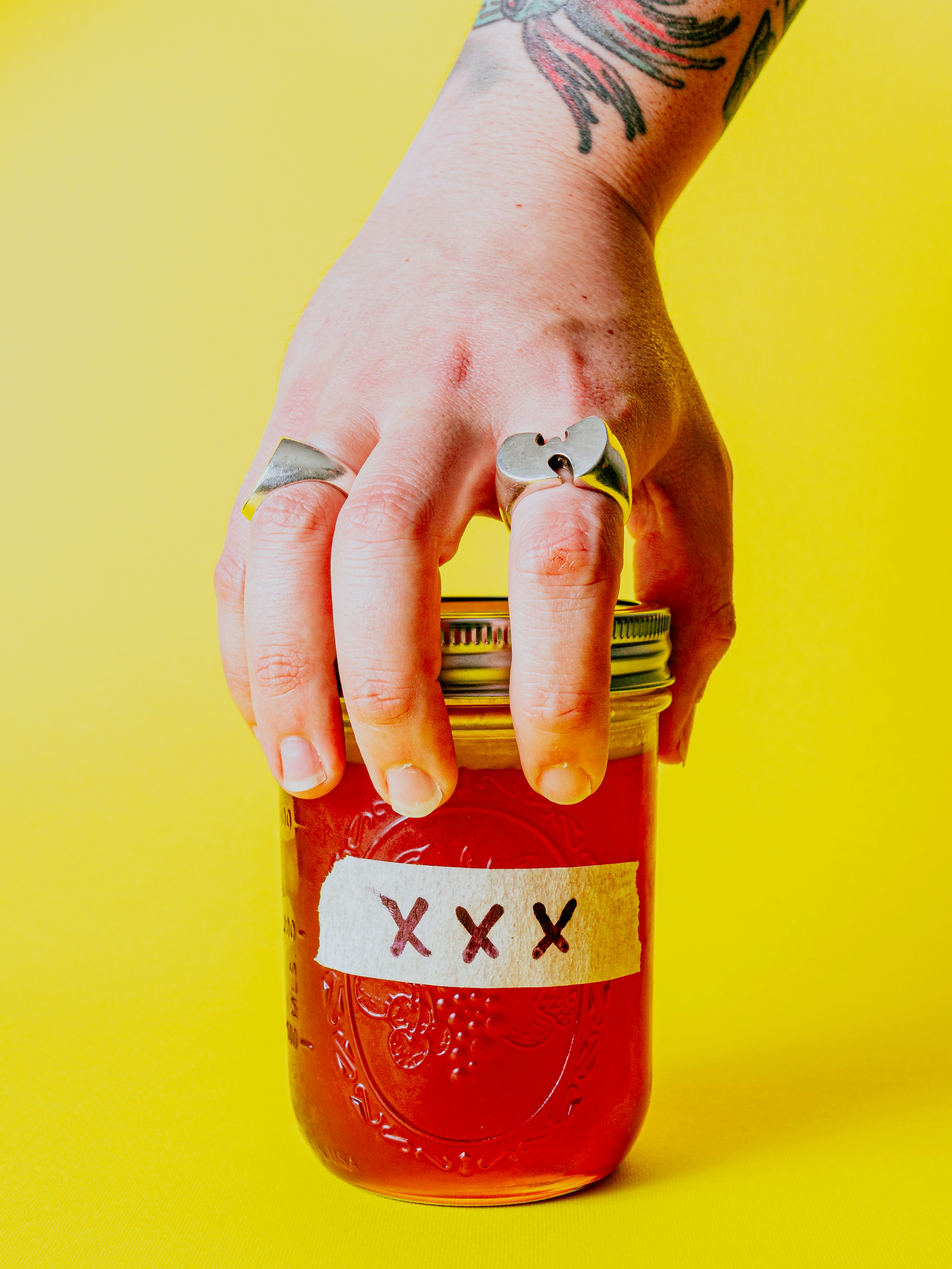 A pale tattooed forearm descends into a bright yellow frame, fingers curled over the gold lid of a Ball mason jar filled with hot honey. Two heavy silver rings sit on the fingers; a strip of masking tape across the jar is marked with three hand-drawn Xs in dark ink. Faint scars are visible across the back of the hand. Clean single-colour backdrop, hard studio light, strong graphic contrast between the red, gold and yellow.