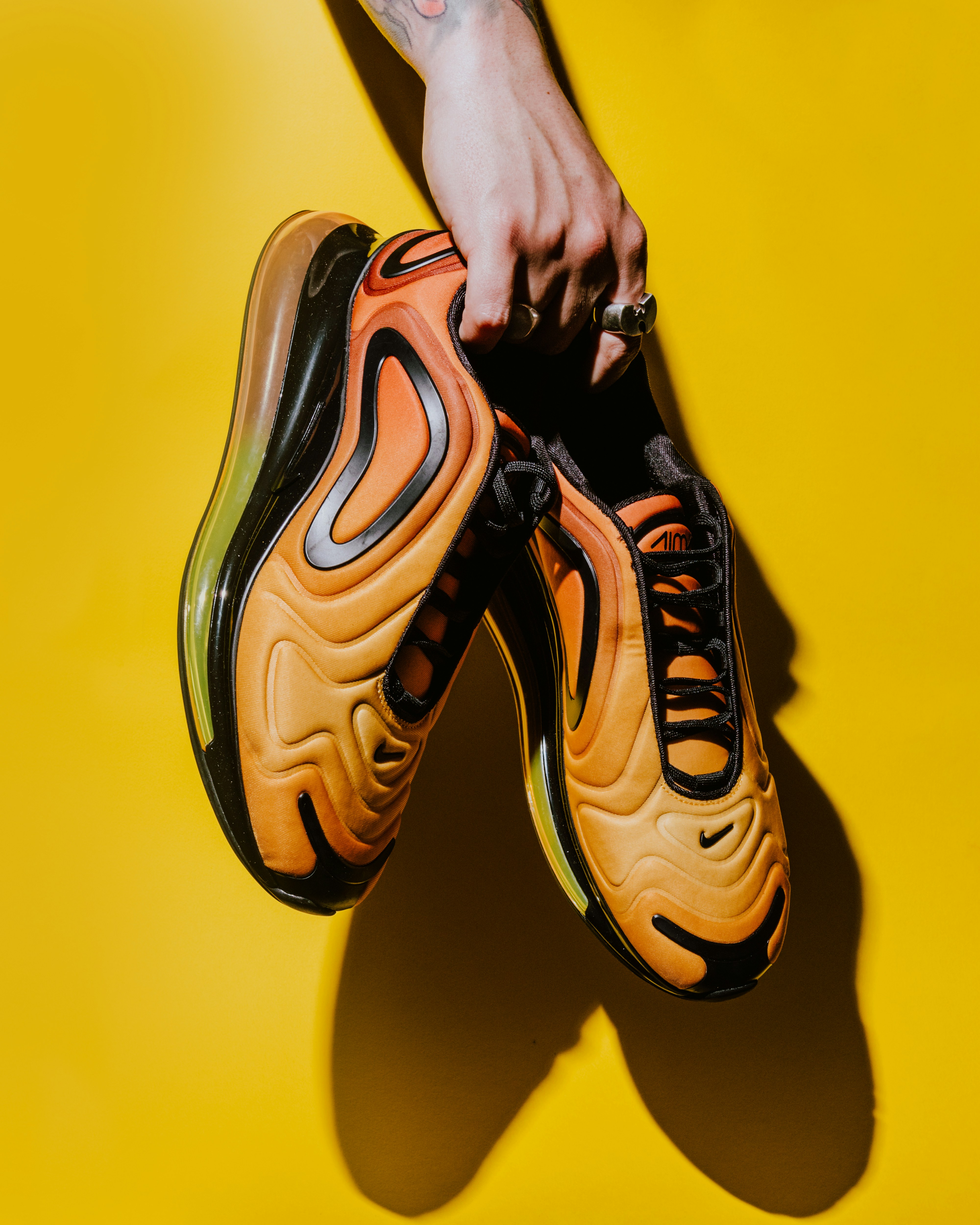 A tattooed hand with two chunky silver rings dangles a pair of Nike Air Max 720 sneakers against a saturated chrome-yellow background. The uppers run from pale peach at the toe through to deep orange, with black swoosh overlays, black laces and a tall translucent air bubble sole tinted orange and lime. The shoes hang side by side at an angle, throwing a long hard-edged shadow down and to the right. Bold, graphic, high-energy colour blocking.