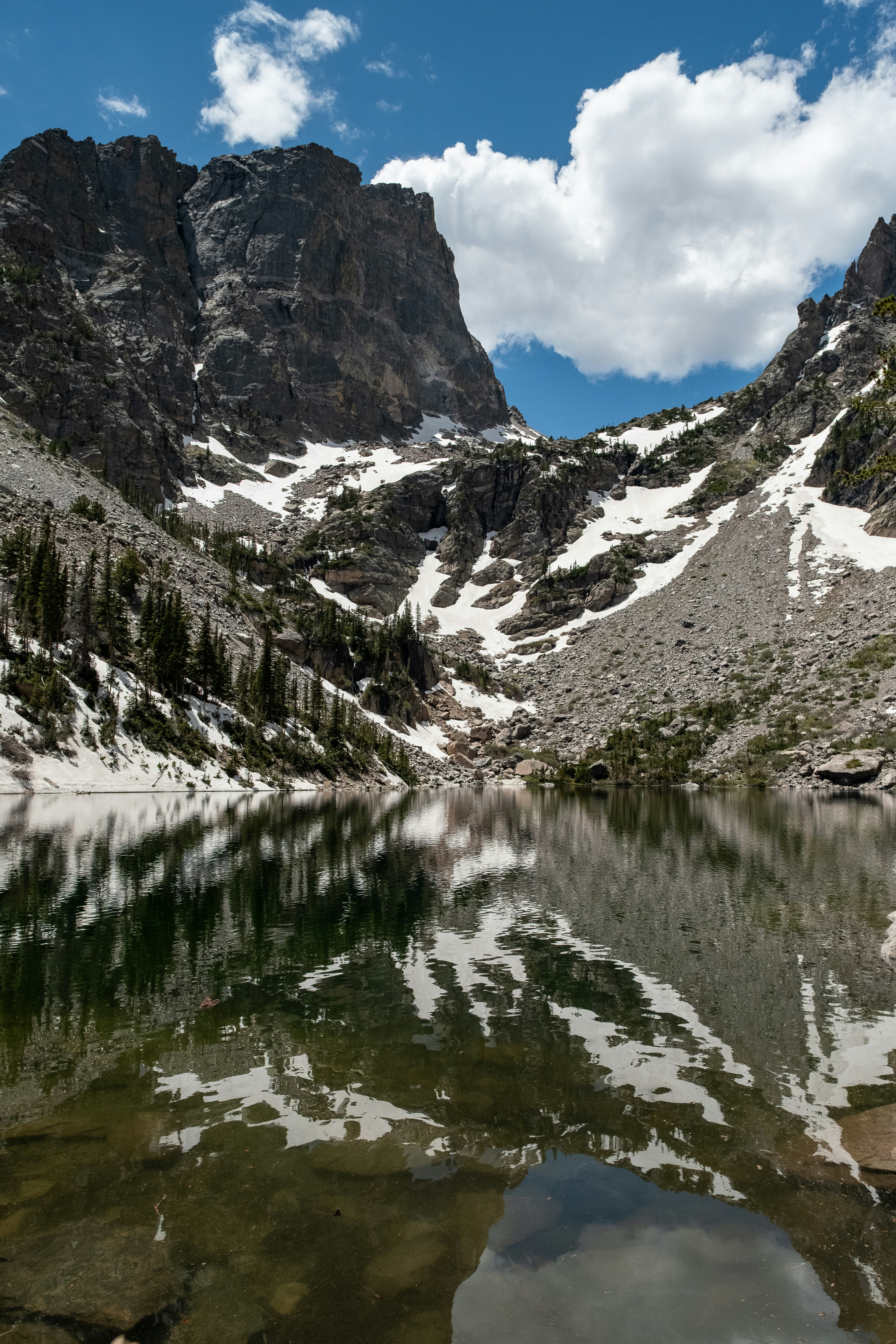 Alpine lake reflects rugged, snow-speckled cliffs under a blue sky; jagged peaks rise above the still water, with evergreen trees along the shore.