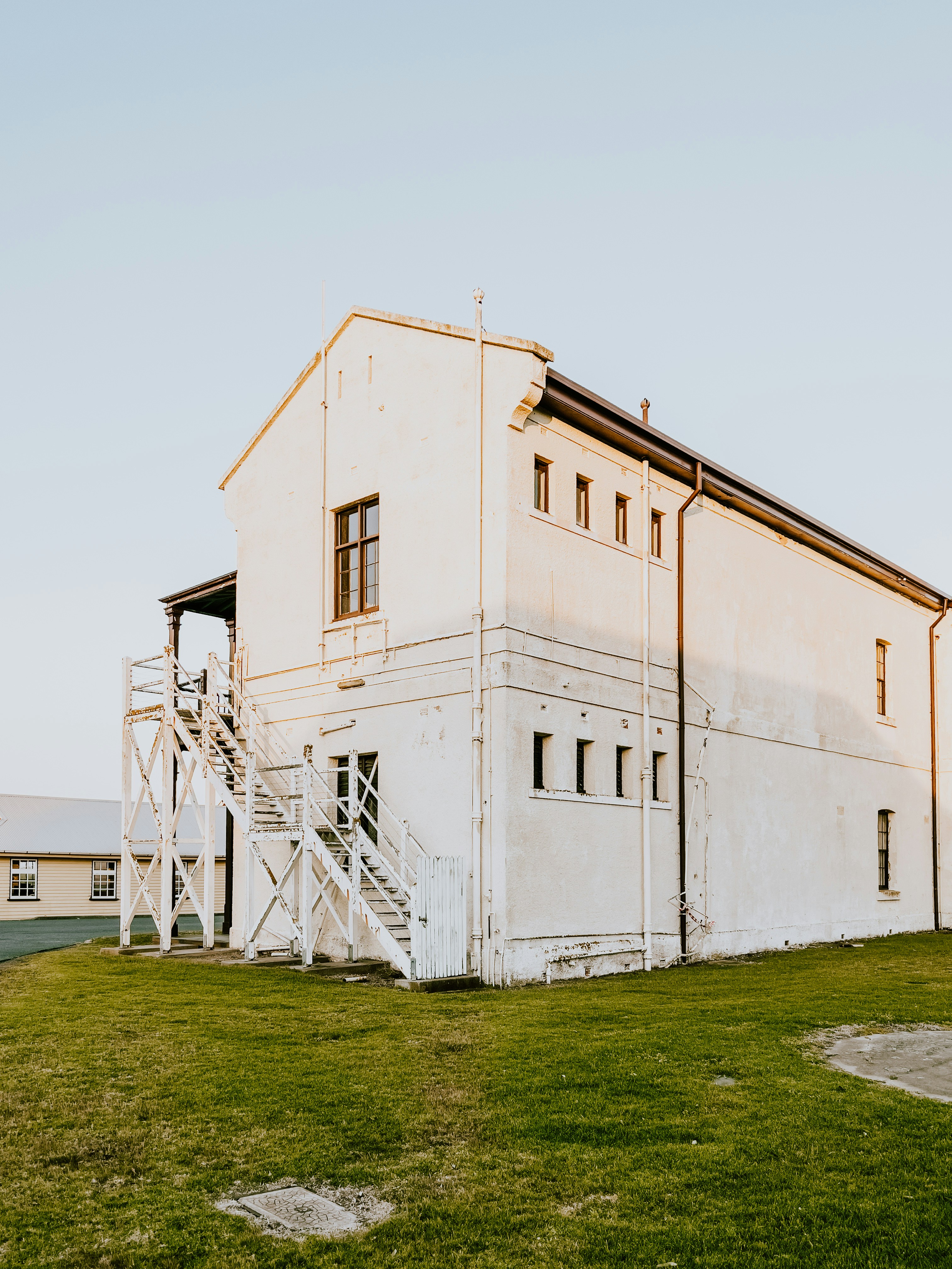 White two-story building with external stairs under a pale blue sky; the structure sits on green grass, with a long shadow and a subtle warm glow.