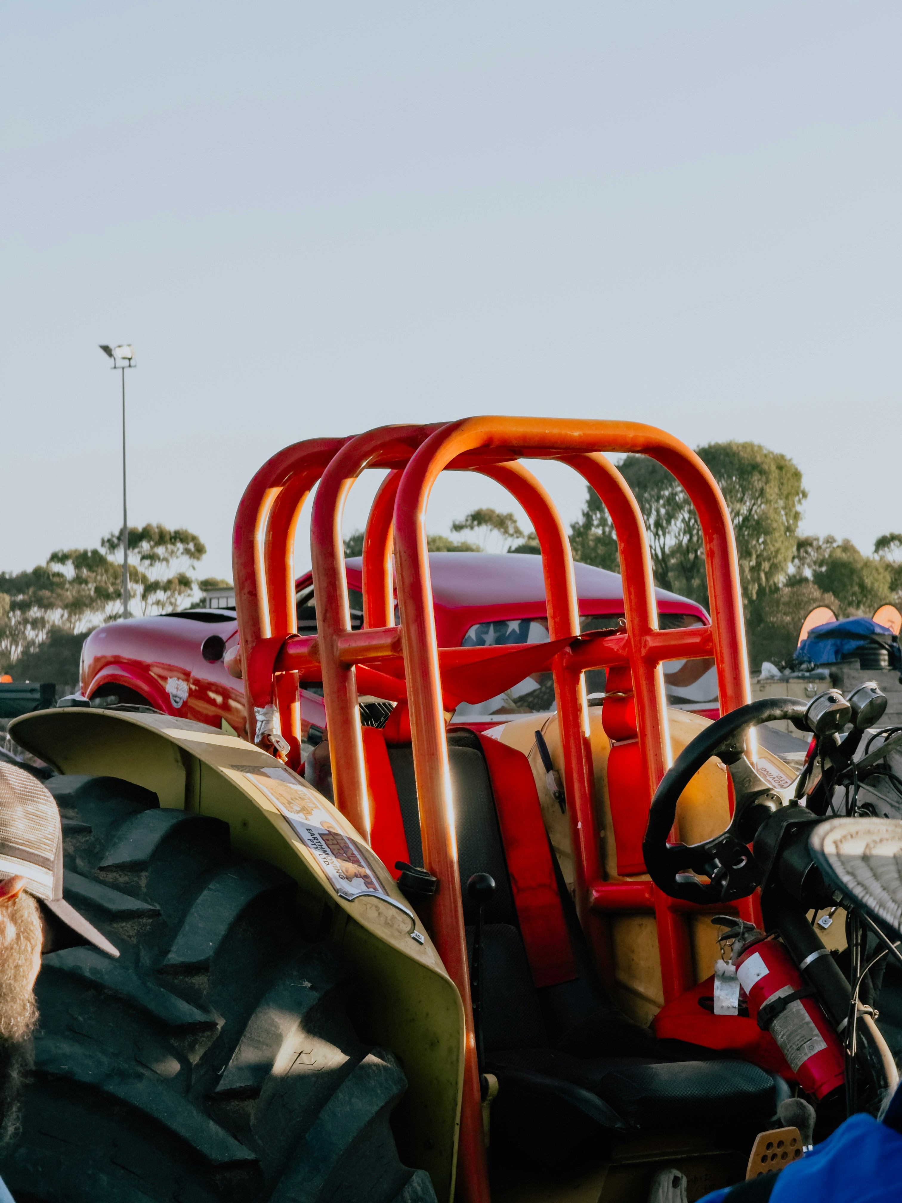 Tight, layered shot at a rural truck and machinery show in late afternoon light. In the foreground, a hulking tractor tyre and yellow mudguard; centre frame, the glowing red-orange tubular roll cage of a modified vehicle with a bench seat, red fire extinguisher and steering wheel; behind it, the deep red body of a monster truck with a stars-and-stripes panel. Gum trees and a floodlight pole against a flat pale sky. Warm rim light on every steel edge.