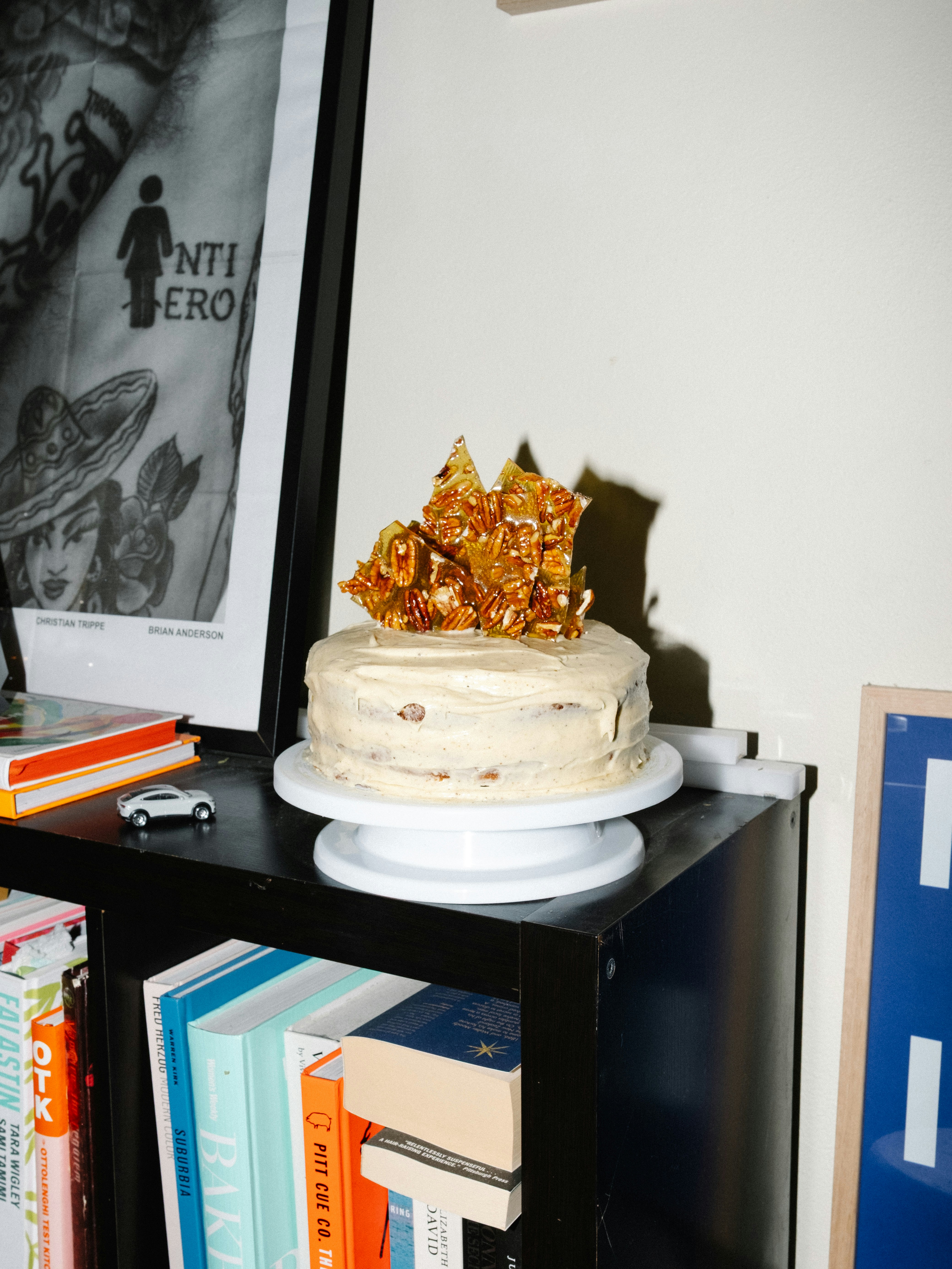 A rustically frosted layer cake on a white plastic turntable, perched on top of a black bookshelf against a plain white wall. Pale cream frosting is swirled with a palette knife; jagged shards of amber pecan praline stand upright on top, casting a hard shadow onto the wall. Framed skate and tattoo-flash posters lean at left, cookbooks and a tiny silver toy car fill the surrounding shelves. Homey, unstyled, warm flash light.