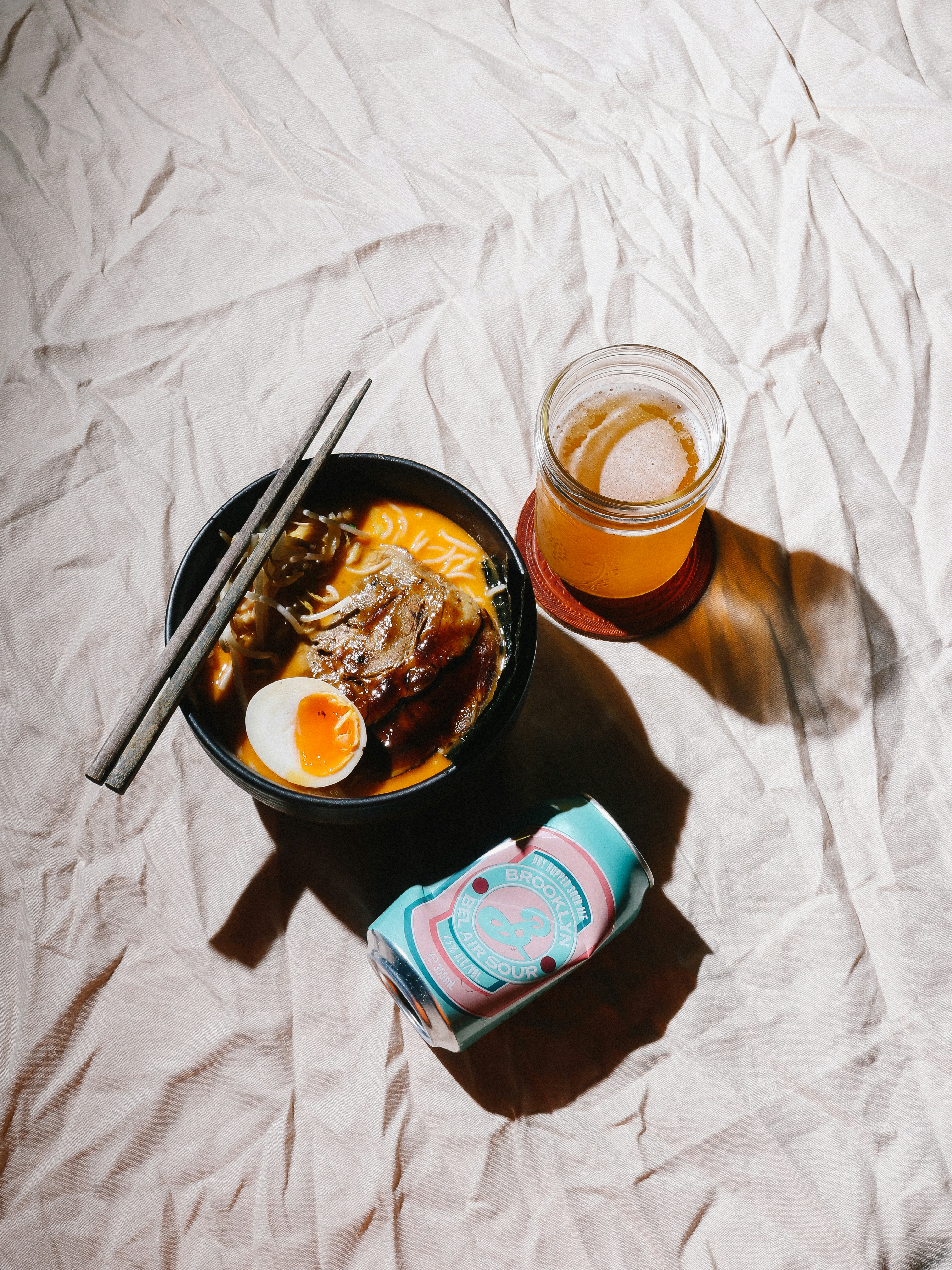A Simple Feast: Ramen Bowl, Soda Can, and Glass of Beer on Wrinkled Linen
