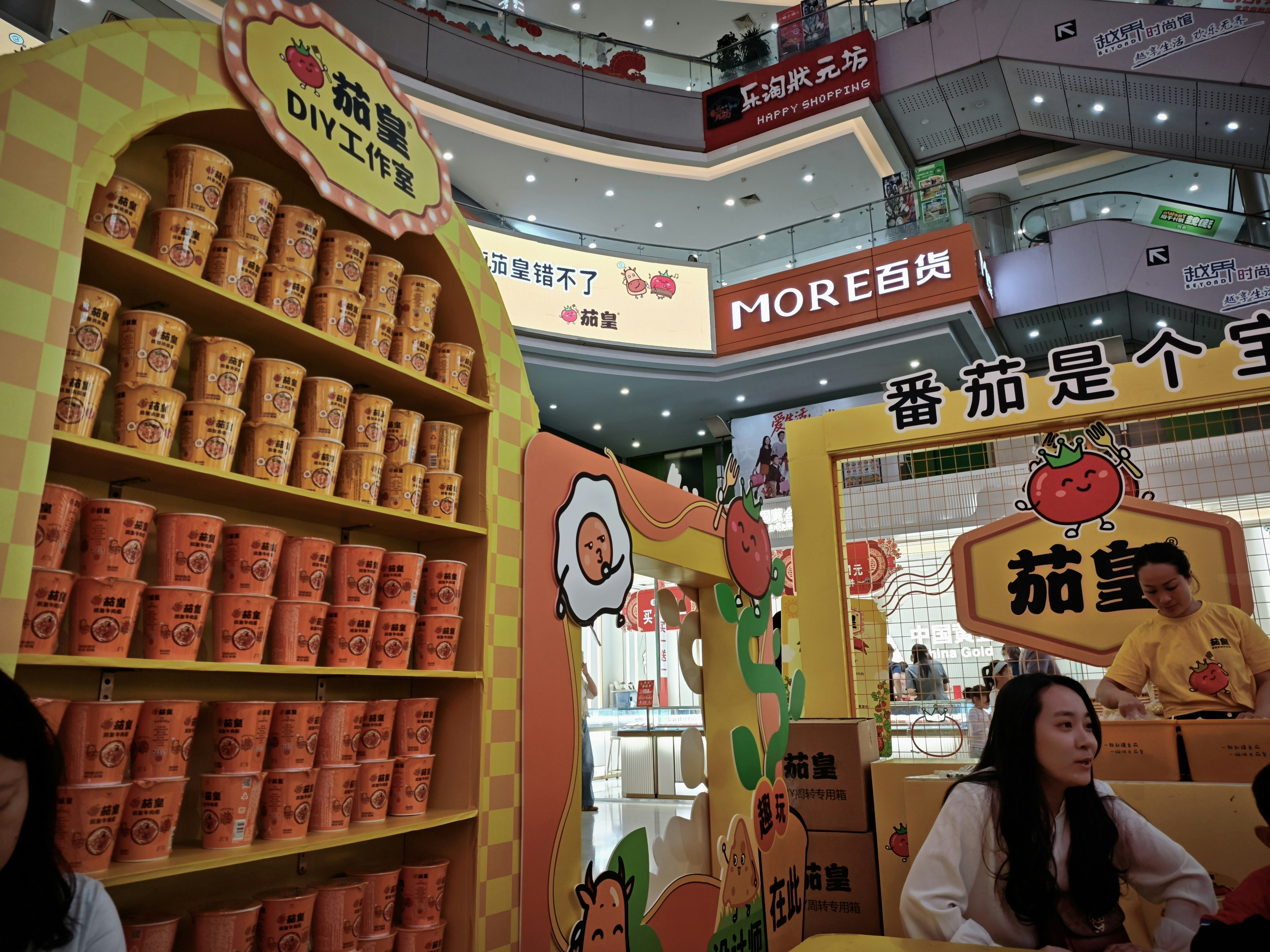 Bright tomato-themed display stands and shelves fill an indoor market area with a shopper in the foreground, highlighting a busy retail setup in a mall. This is a photo of a promotional booth featuring cartoon tomato characters and product boxes.