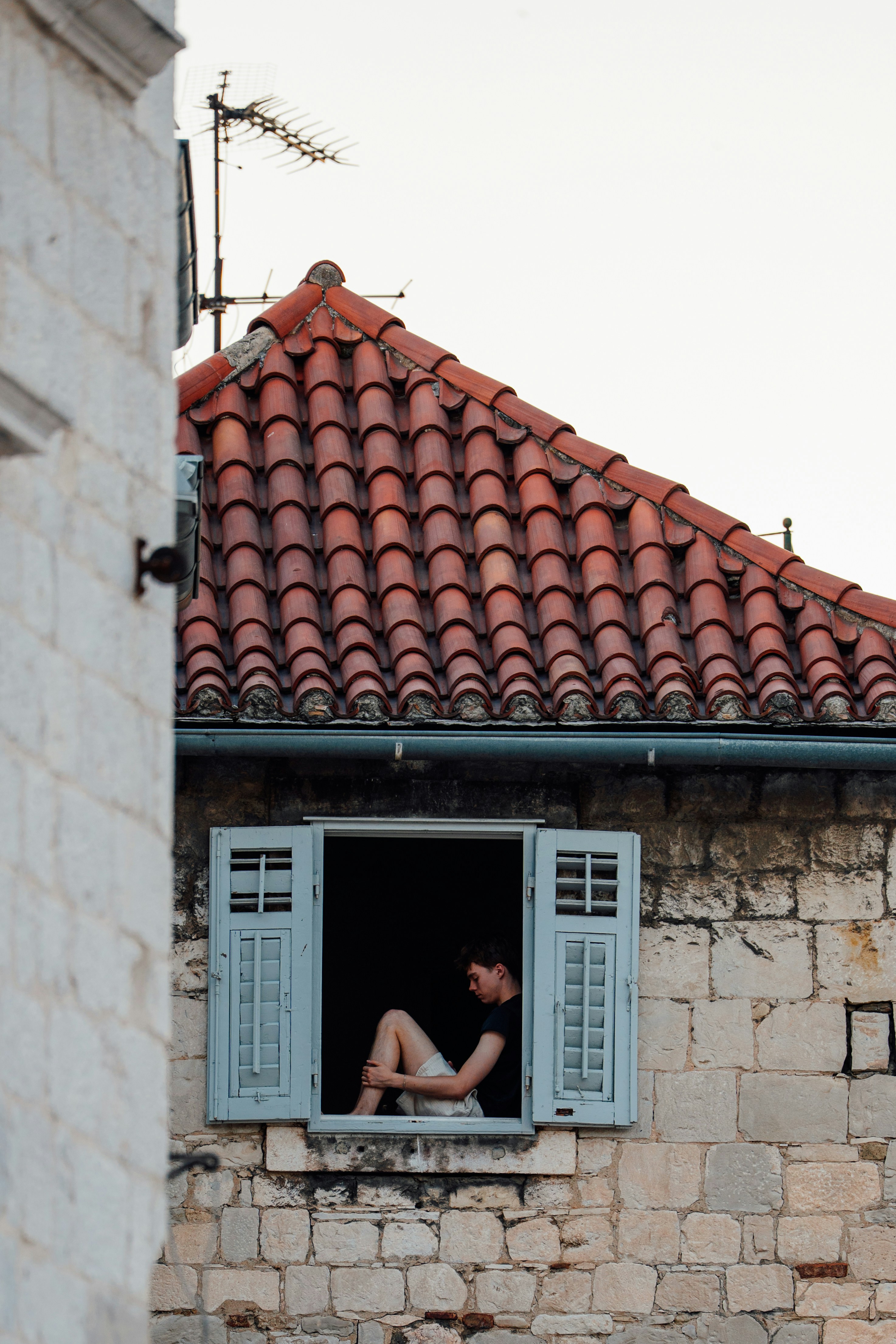A person sits curled in a blue window frame of an old stone building, absorbed in a private moment; the scene is framed by a rustic roof and plastered walls. This is a photographic capture, not a drawing.