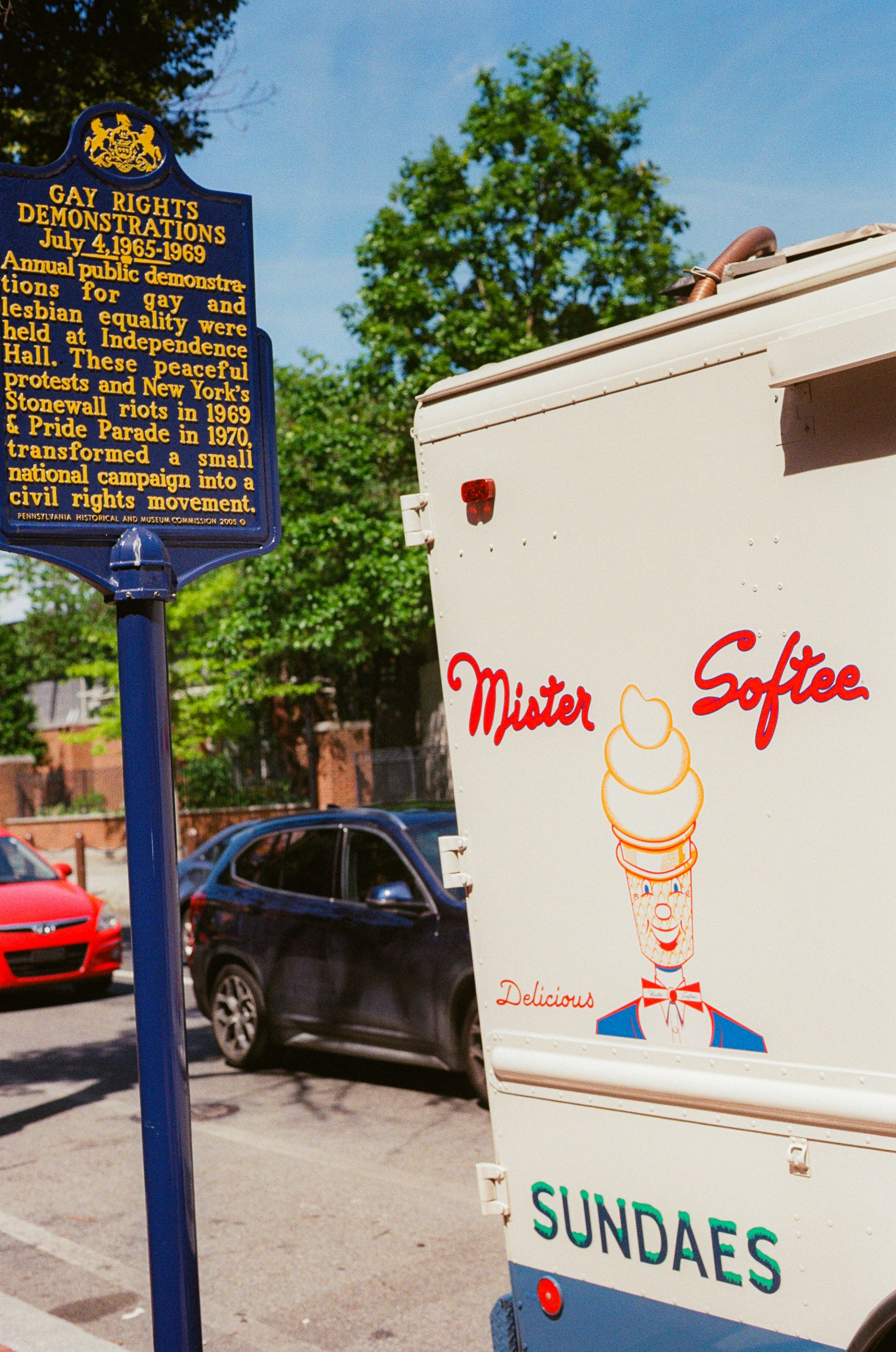 Ice cream truck with graphic cone art sits beside a vintage historic marker on a sunlit urban street.