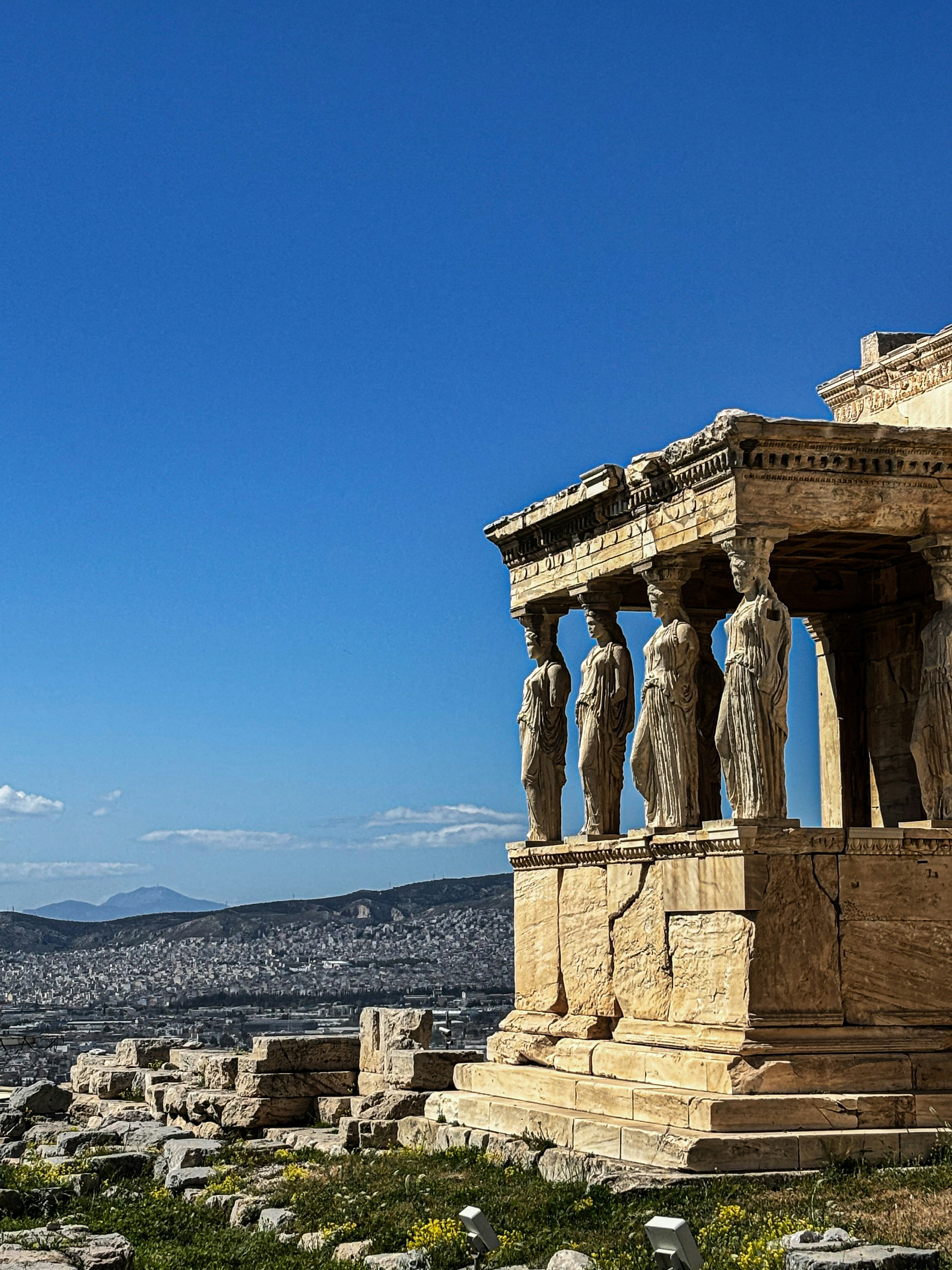 Ancient temple ruin with life-sized statues along a stone wall under a clear blue sky; rugged hillside and distant cityscape visible. (Note: this is a historical structure photo.)