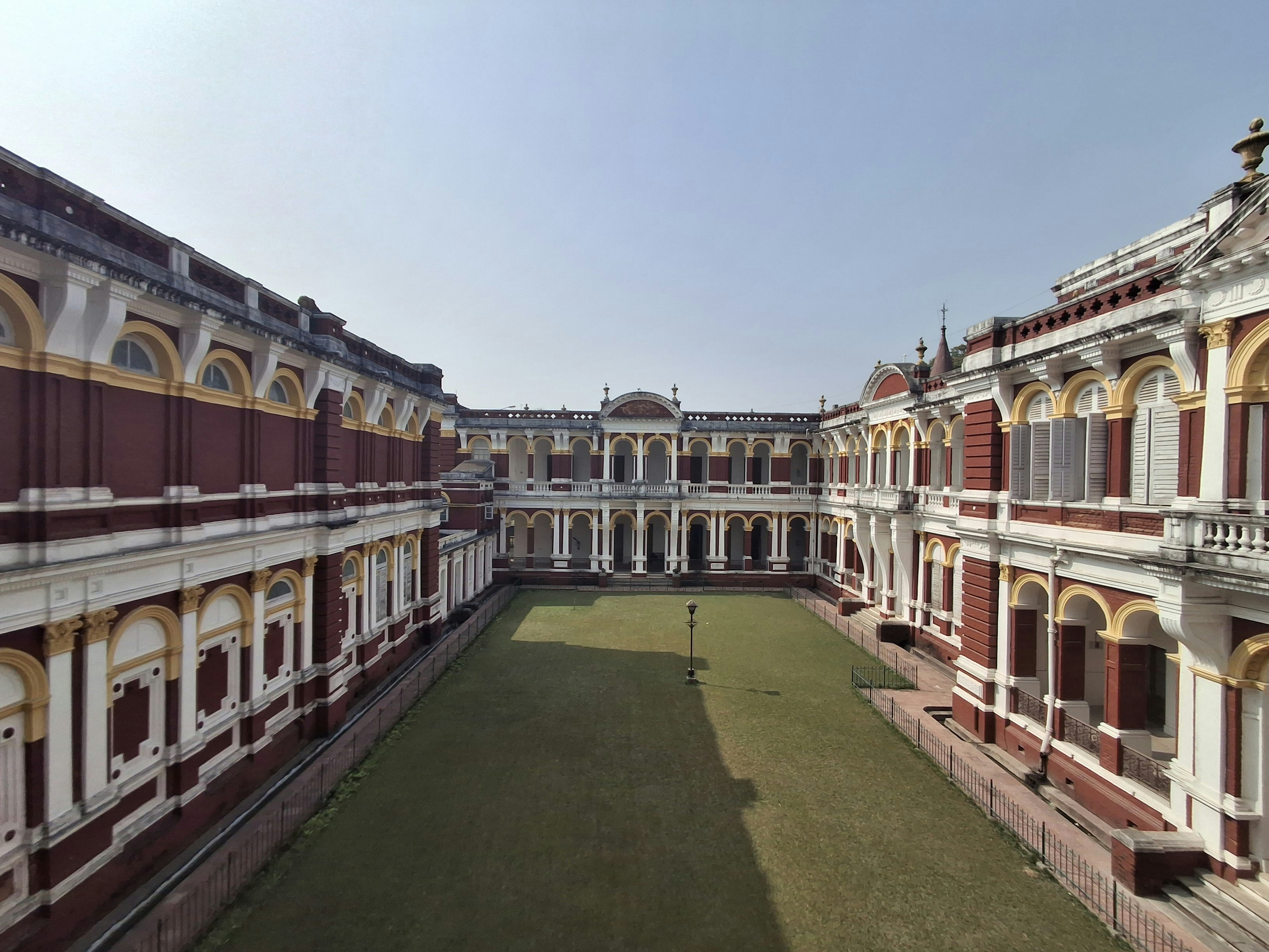 An expansive classical courtyard framed by red and cream arcaded walls under a clear blue sky, with a central grassy strip leading to a distant building; a calm architectural scene captures depth and symmetry.